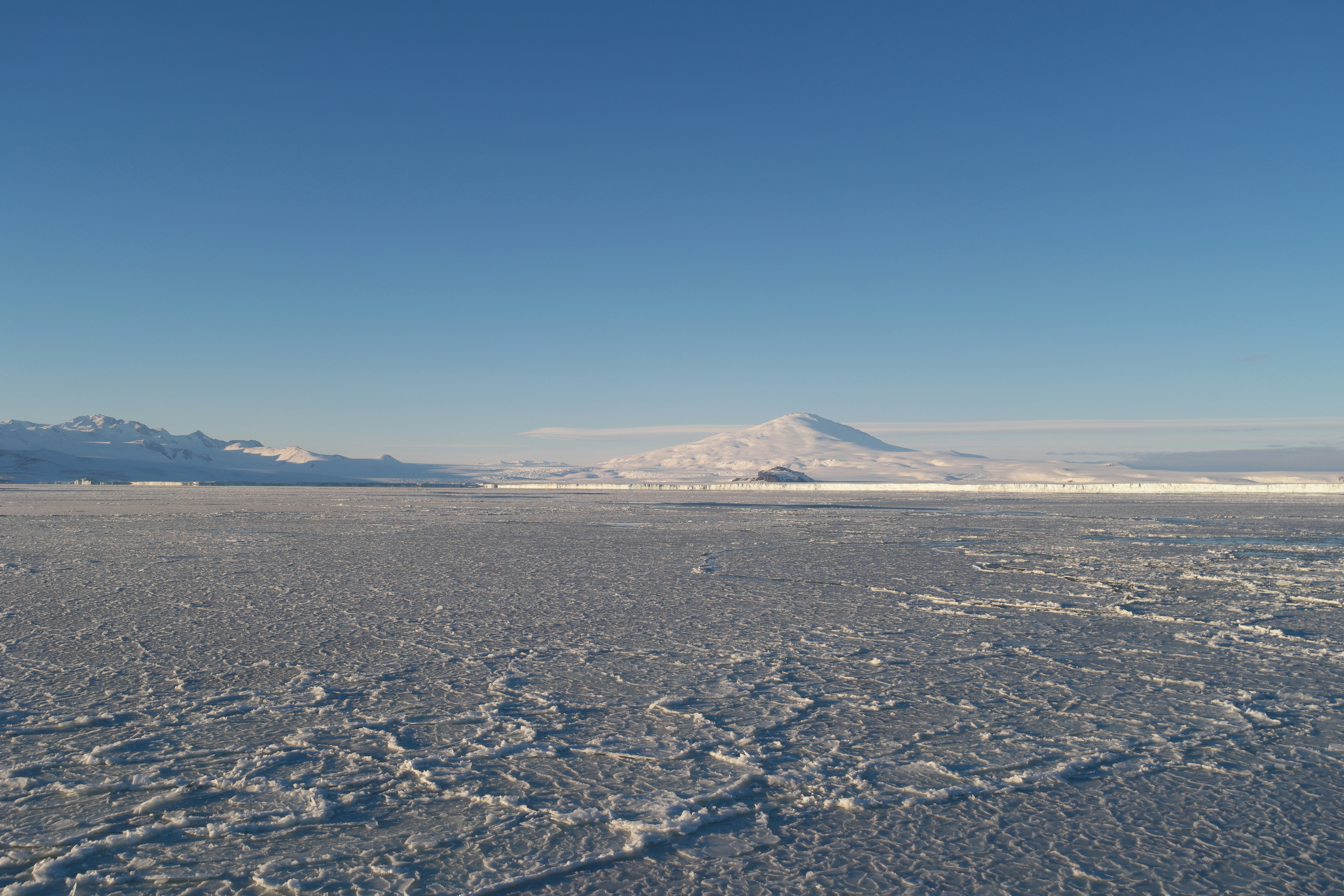 Vast icy landscape with a distant snow-capped mountain under a clear blue sky. The scene captures the serene beauty of a frozen expanse.