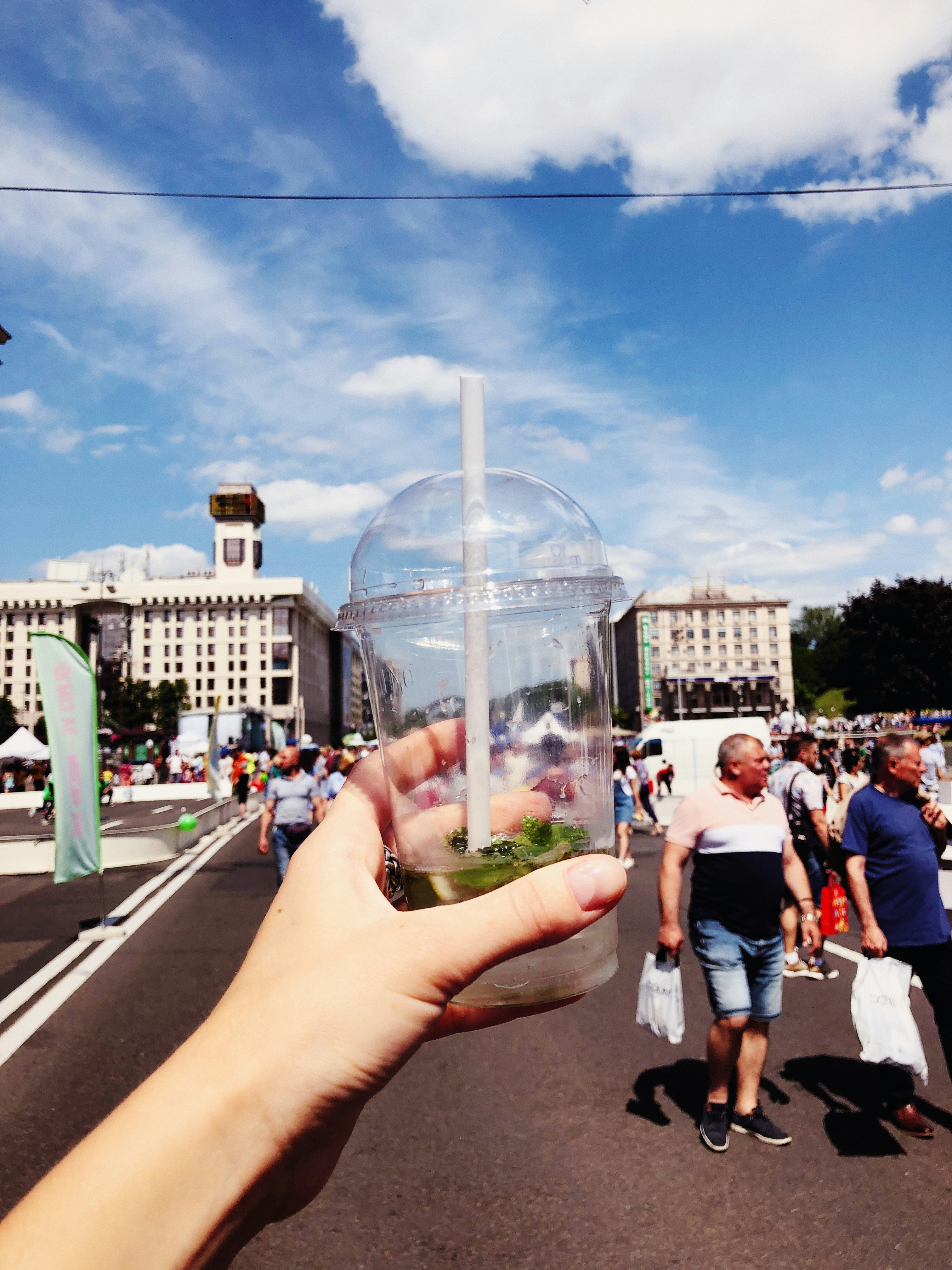 A clear plastic cup with a straw is held up in a busy city square, crowd and buildings in the background; a bright day with blue sky and scattered clouds. The image appears to be a candid street scene captured outdoors.