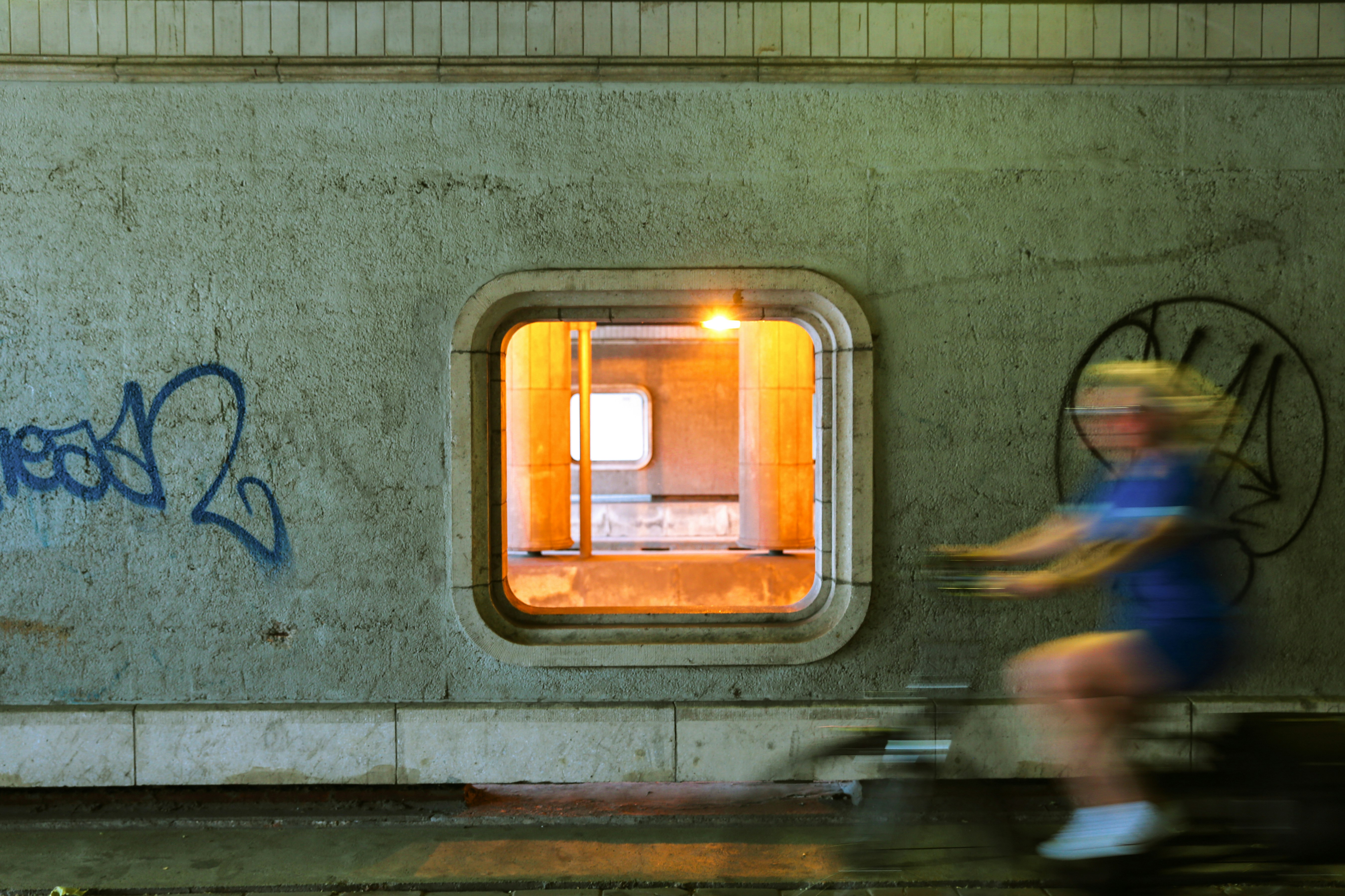 Blurred cyclist passes by a graffiti-marked concrete wall with a glowing window.