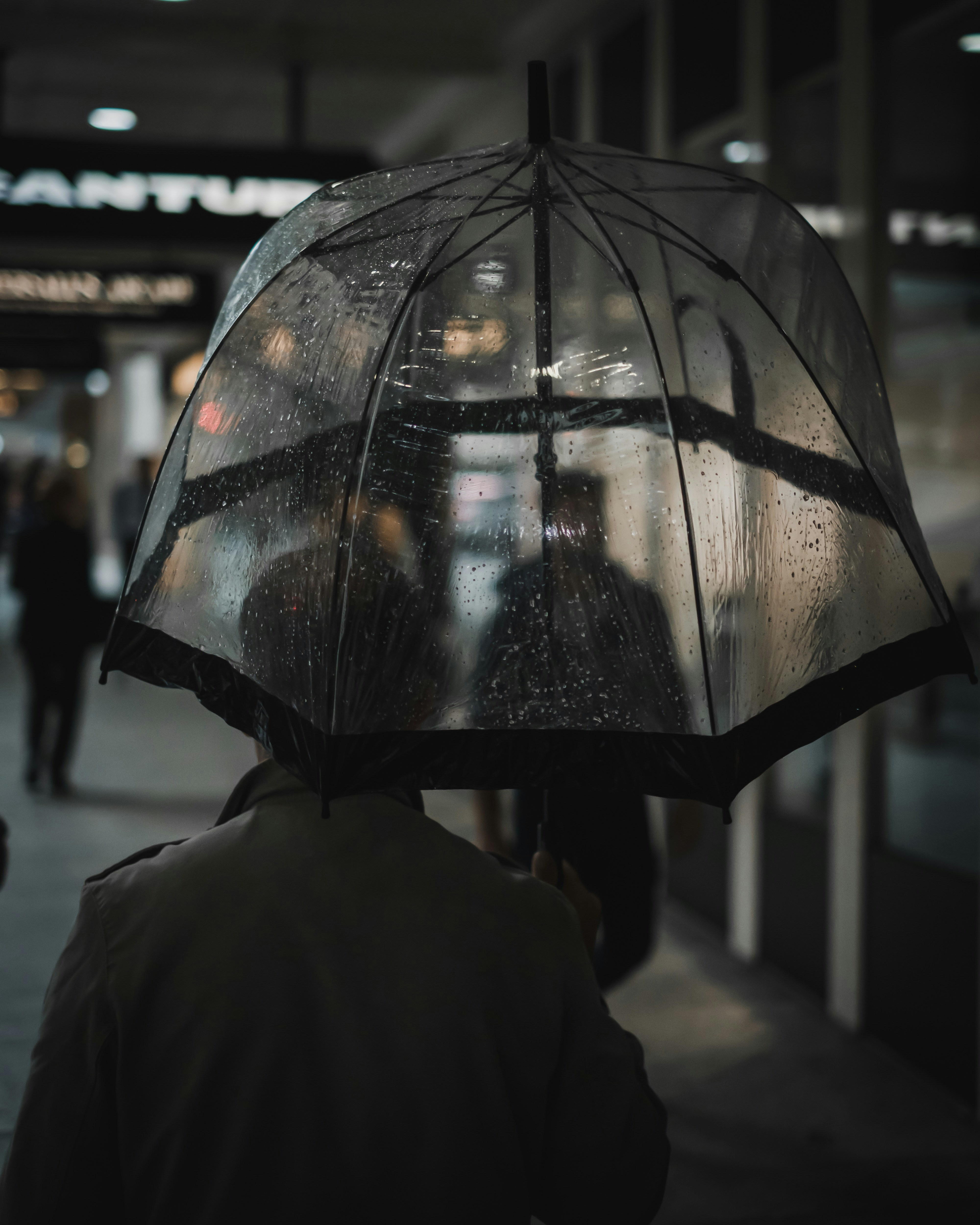 A figure holding a transparent umbrella, surrounded by blurred silhouettes in a rain-soaked urban setting. The reflections and raindrops create a moody atmosphere.
