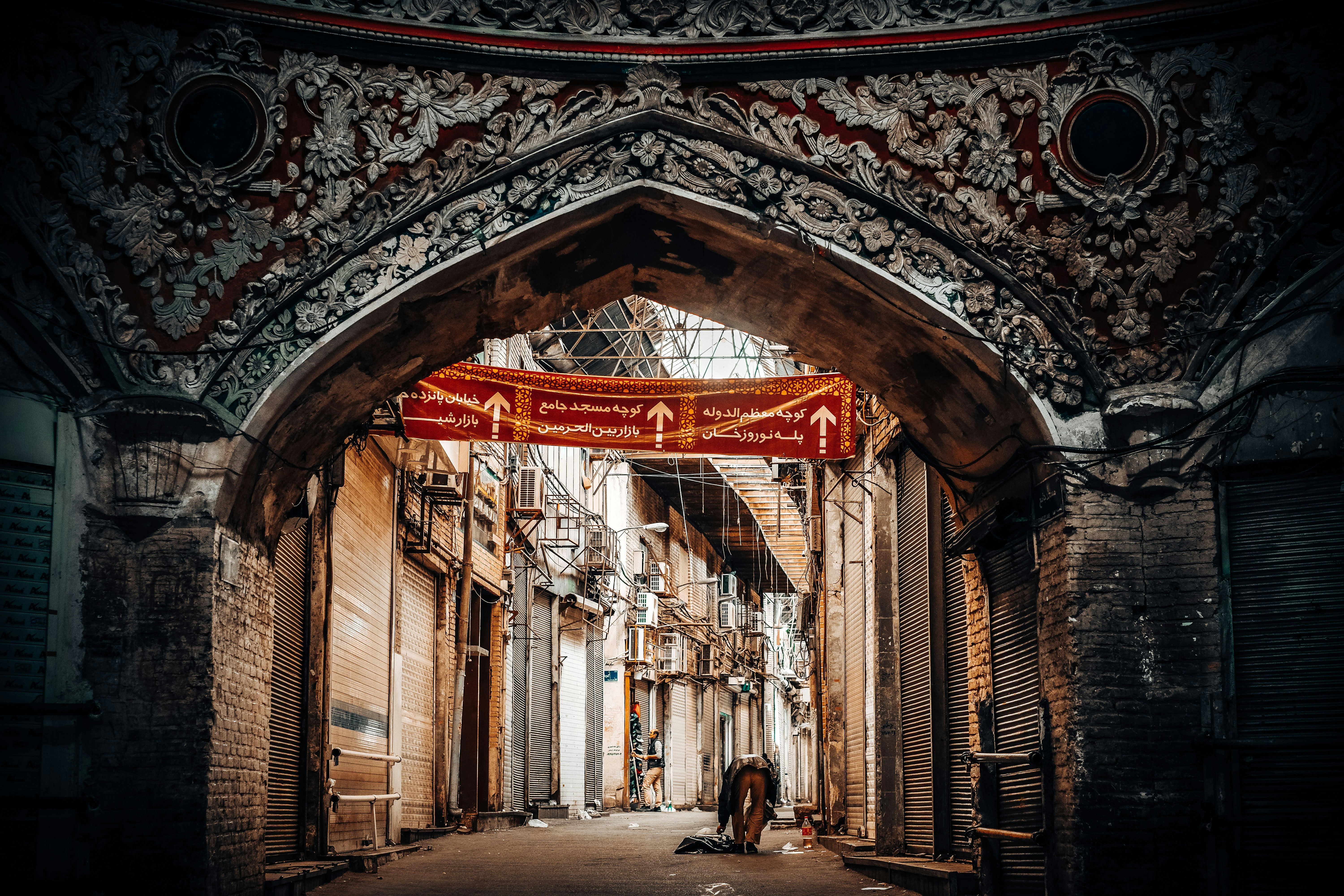 Ornate archway leading into a quiet, narrow bazaar with closed storefronts and a person sweeping the ground.