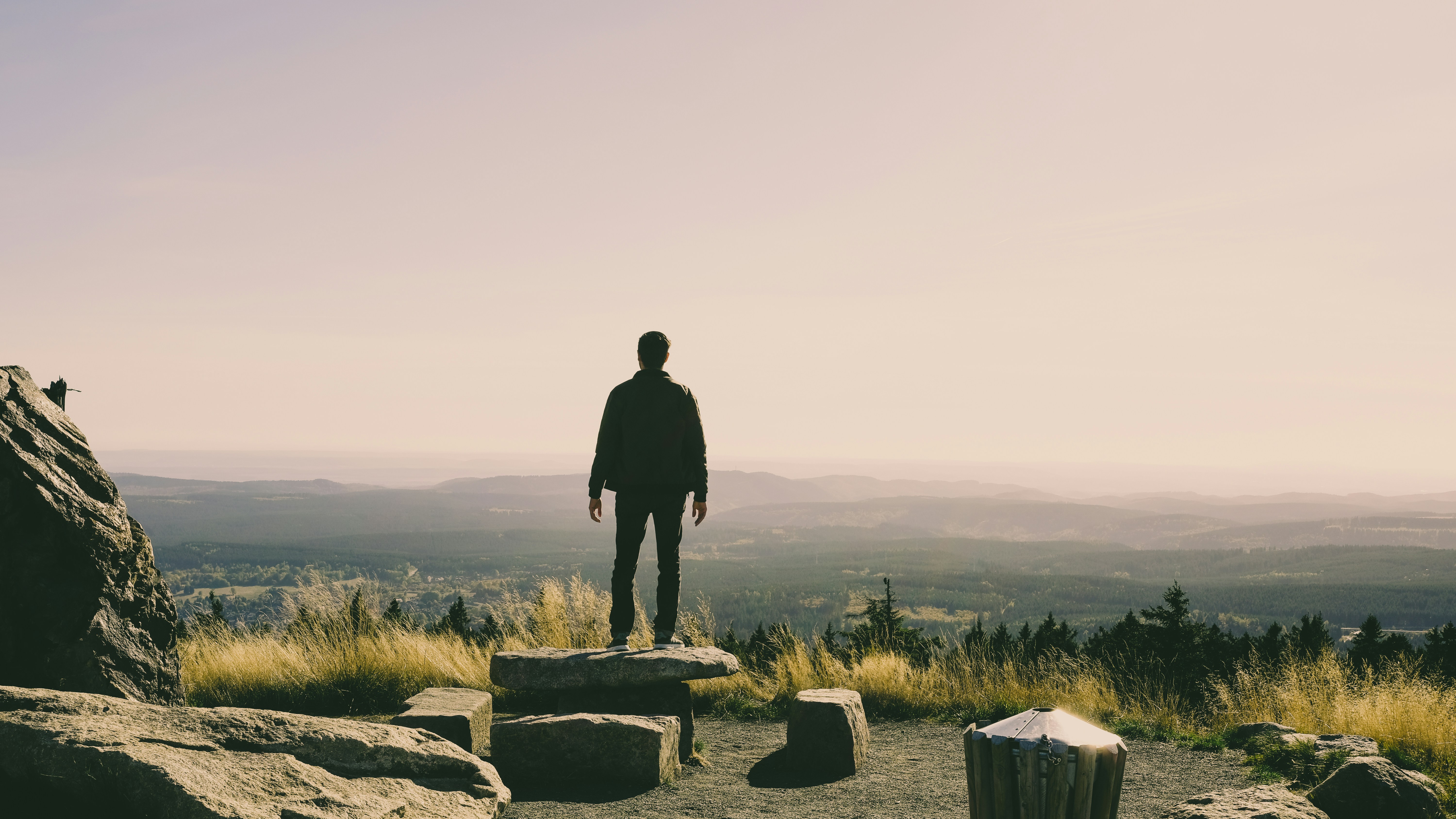 Silhouette of a lone figure standing atop a rocky outcrop, overlooking a vast landscape under a soft pastel sky.