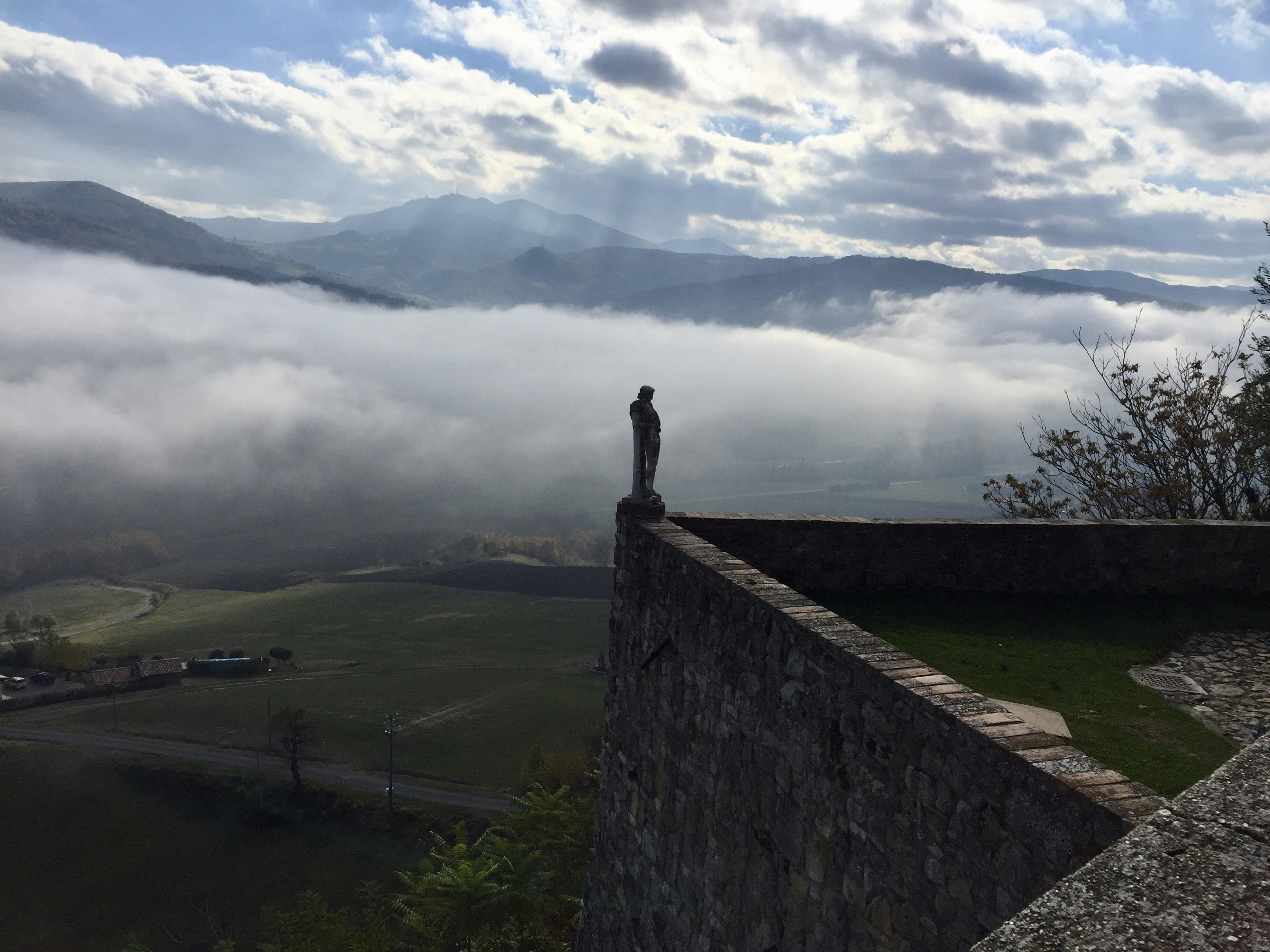 Figure standing on a stone wall overlooking a fog-covered valley with mountains in the distance.