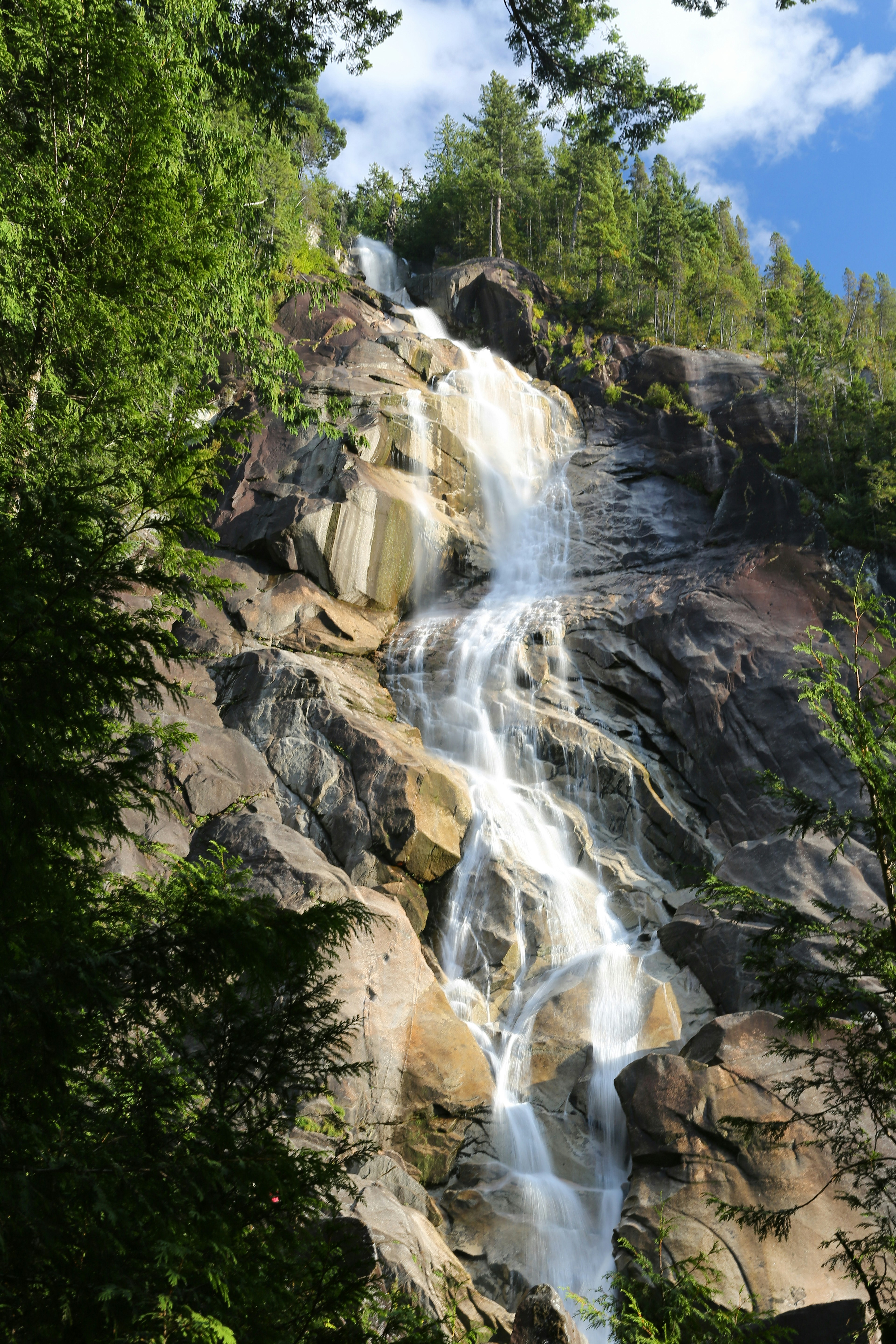 A majestic waterfall gracefully descends rocky cliffs, surrounded by lush greenery and a clear blue sky.