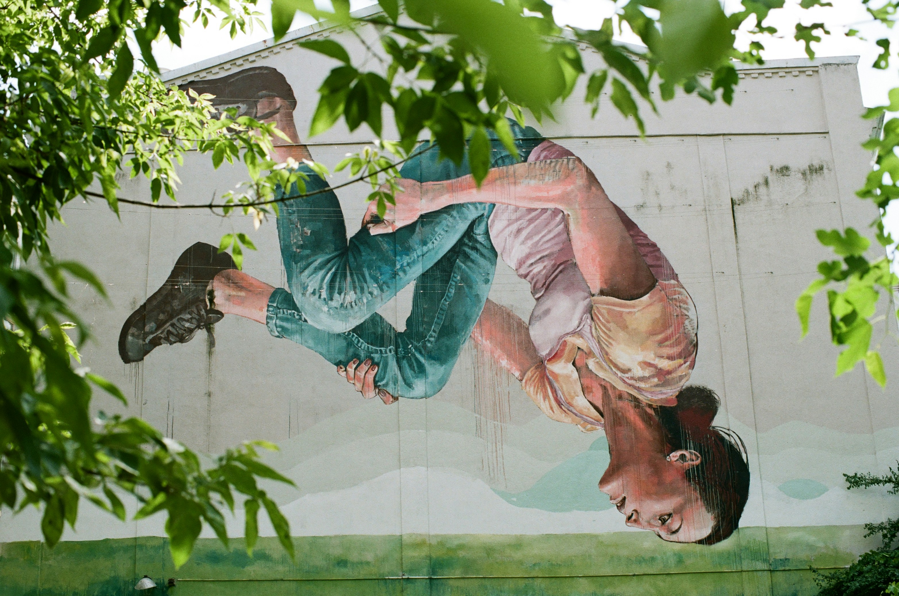 Mural painting of a person hanging upside down on a concrete wall, framed by leafy branches.