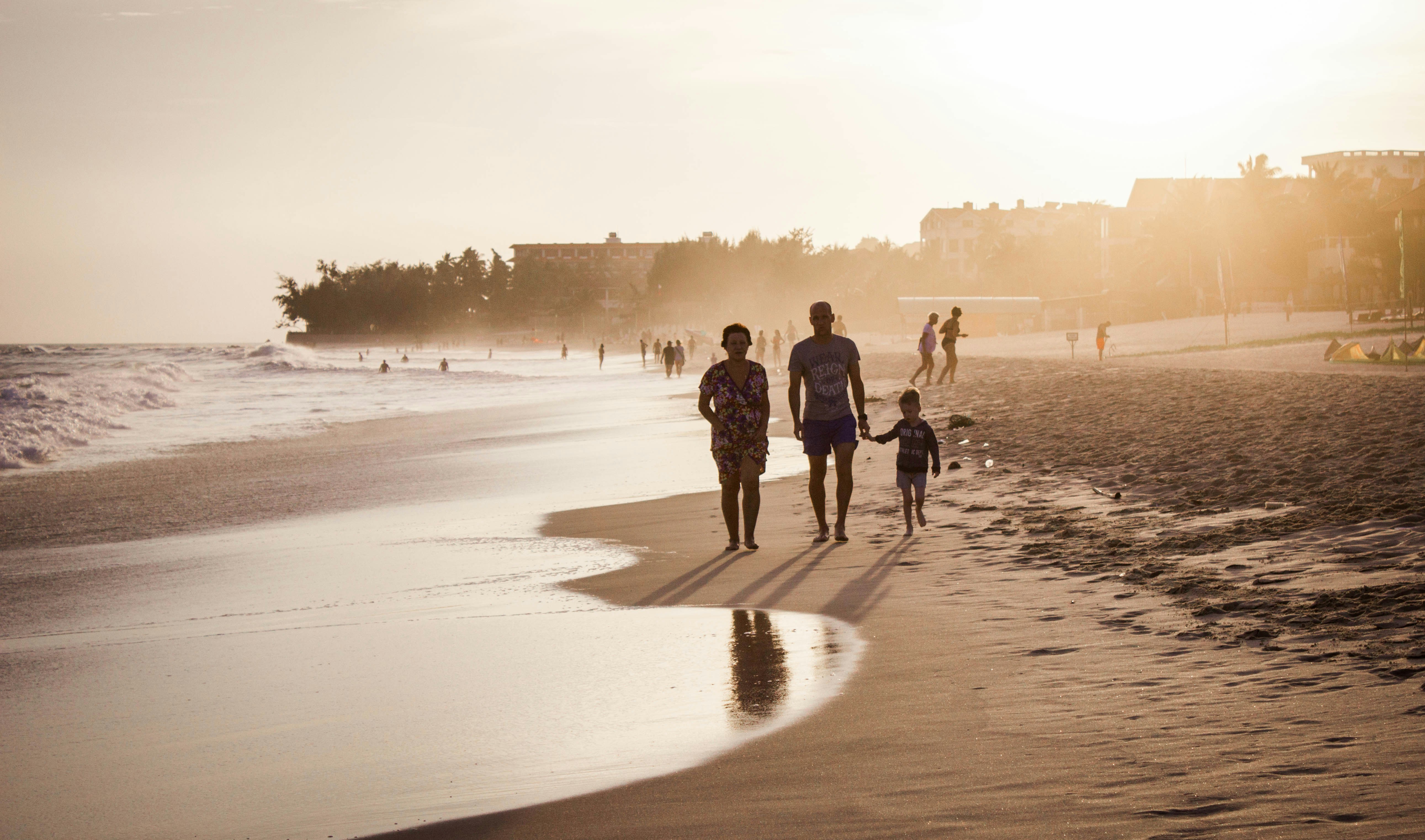 Family walking hand in hand along a sunlit beach at dusk, with gentle waves lapping at their feet.