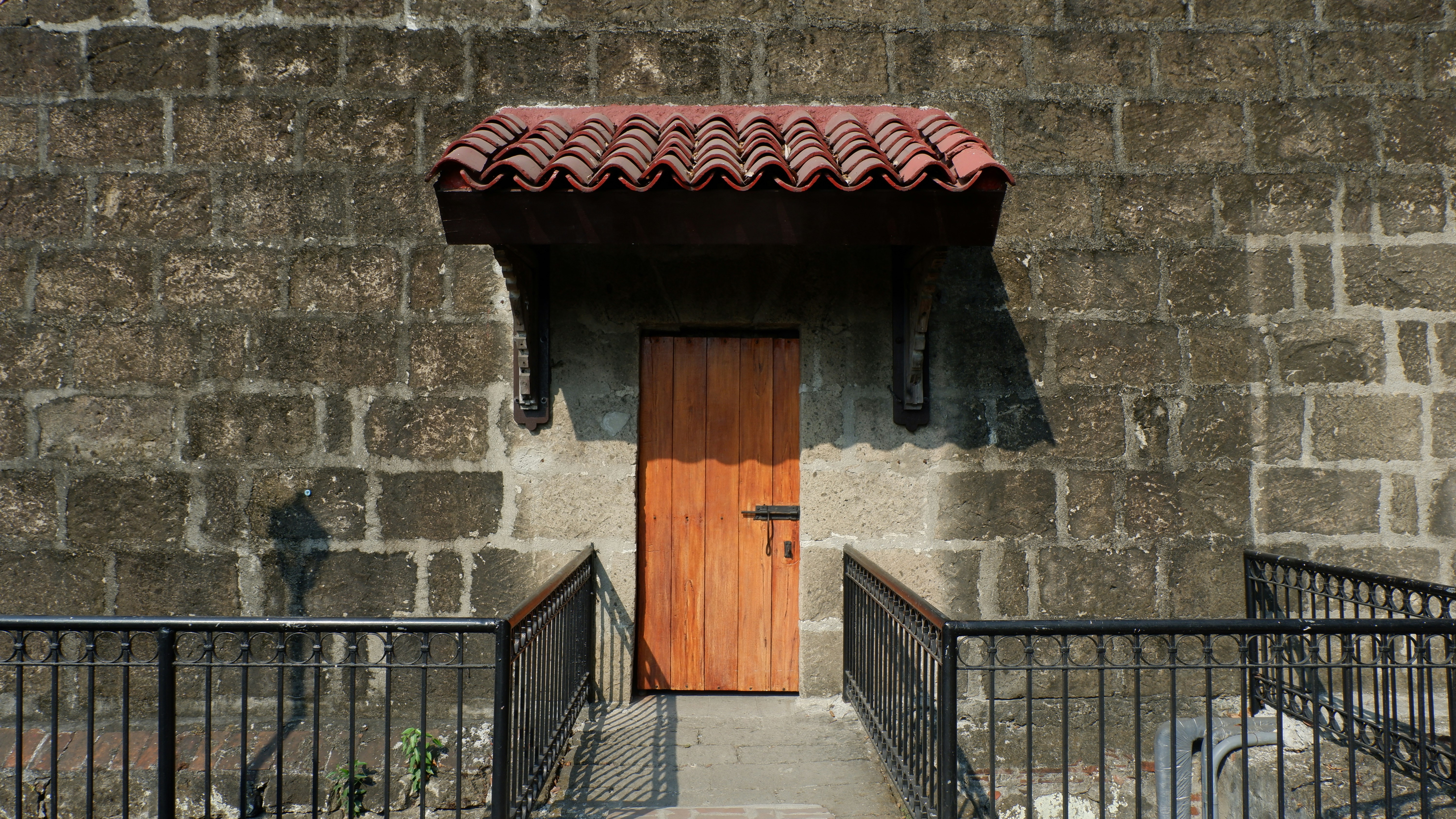 A wooden door framed by a textured stone wall and topped with a traditional red-tiled roof, leading to an unknown interior.