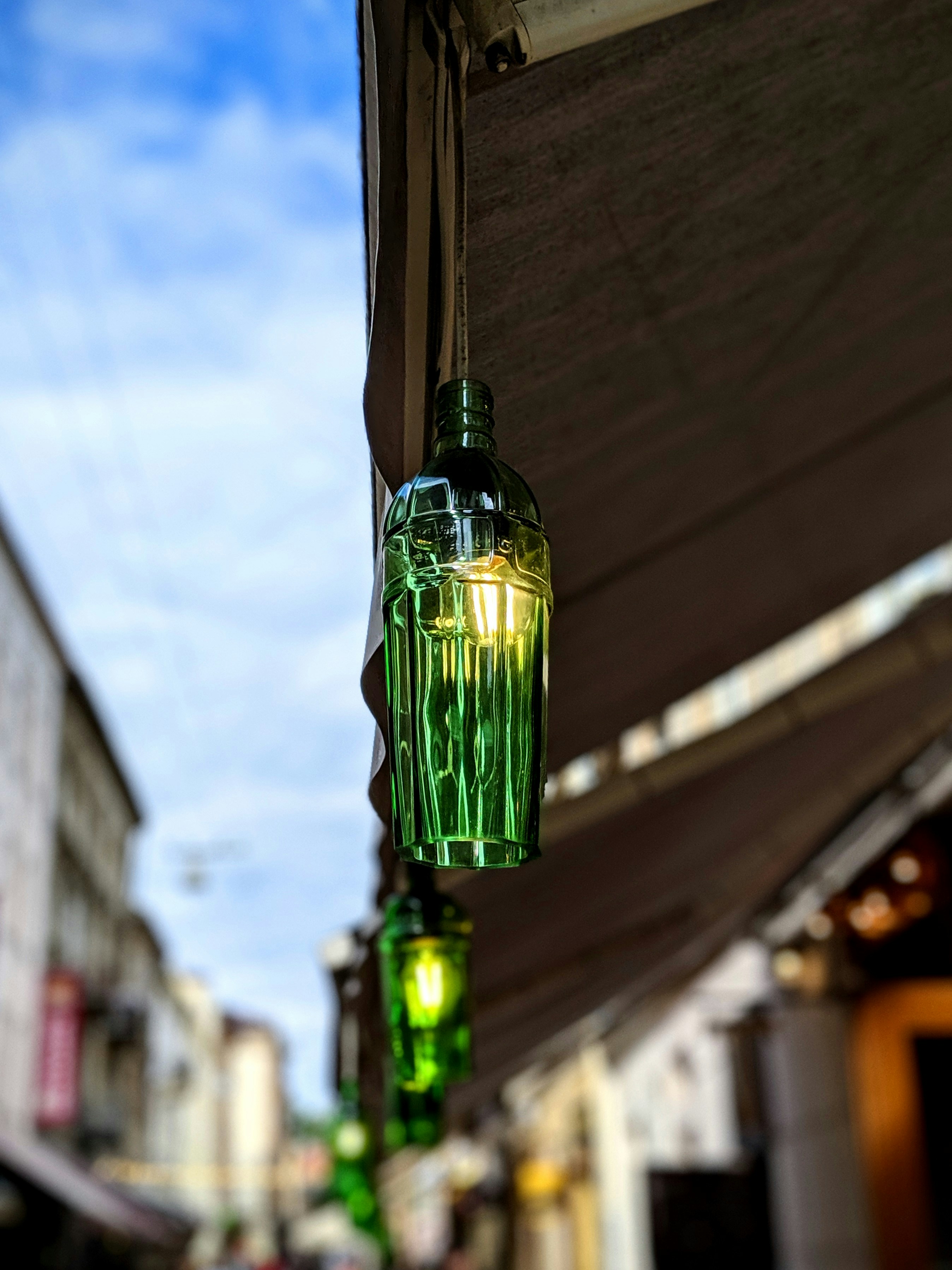 Green glass bottle lanterns hang in a sunlit city alley, with a shallow depth of field blurring the street behind.