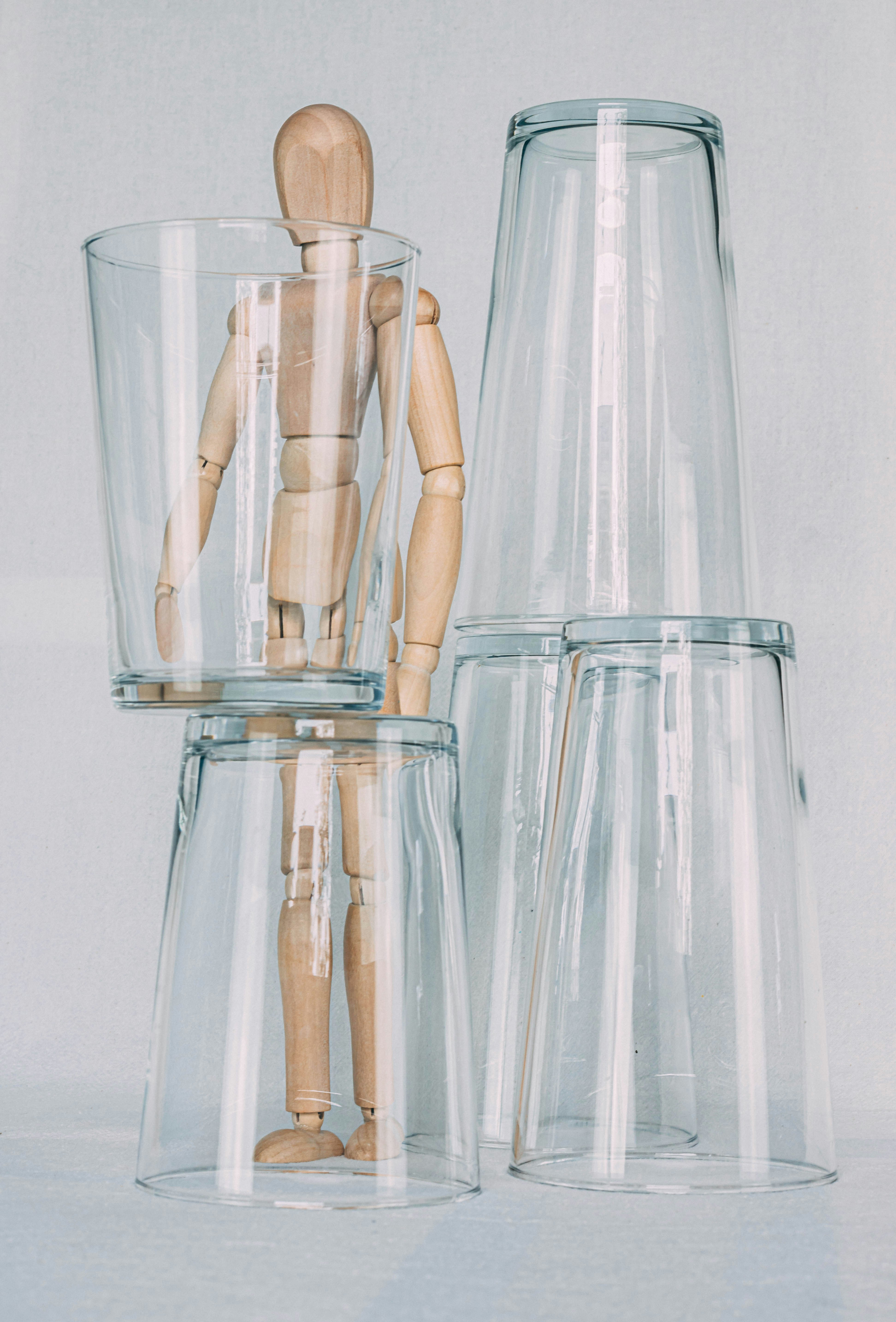 Still-life photograph of a wooden mannequin nestled inside stacked glass tumblers against a pale backdrop.