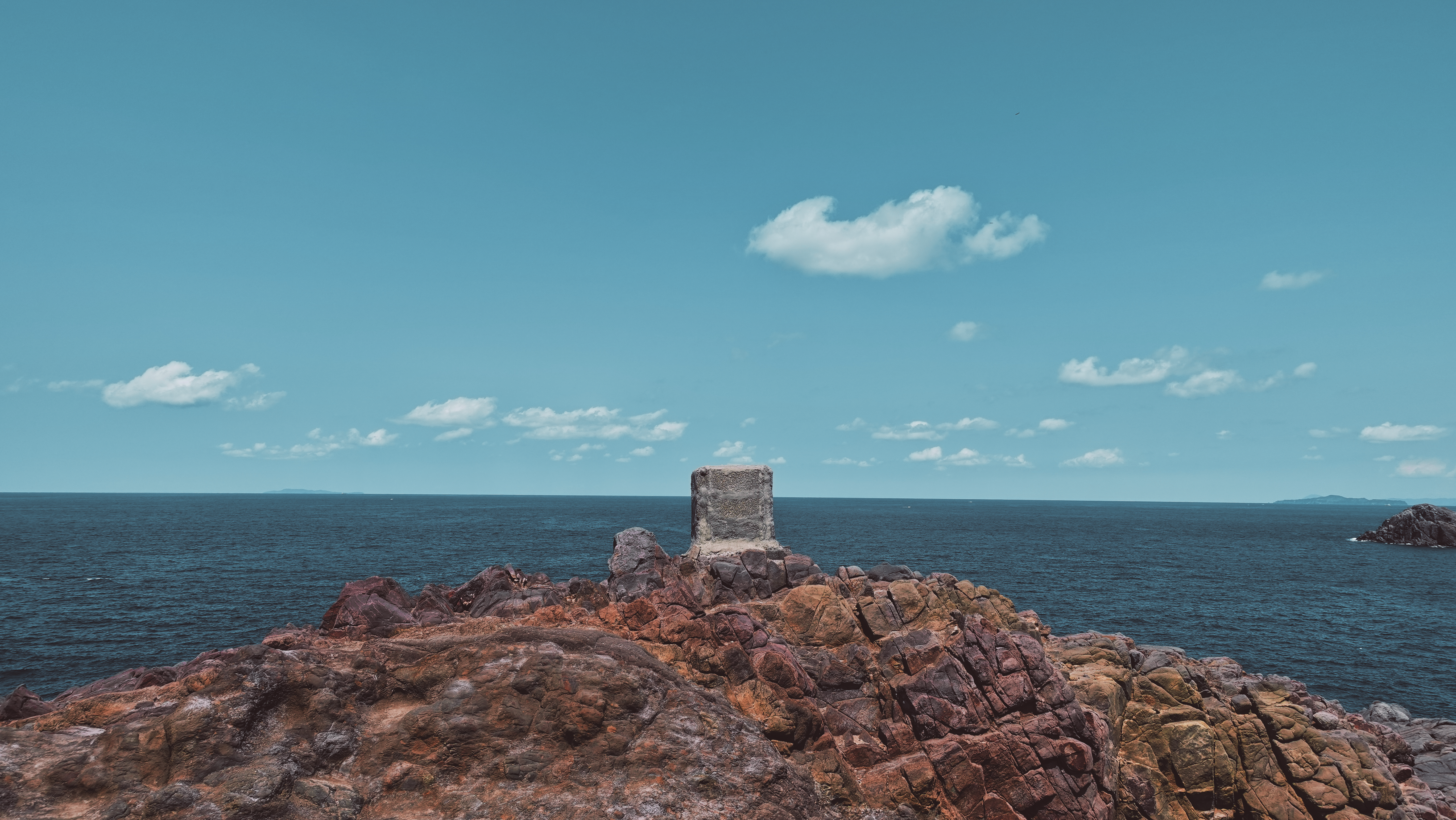 A solitary stone structure stands atop a rugged cliff, overlooking the vast ocean under a clear blue sky.