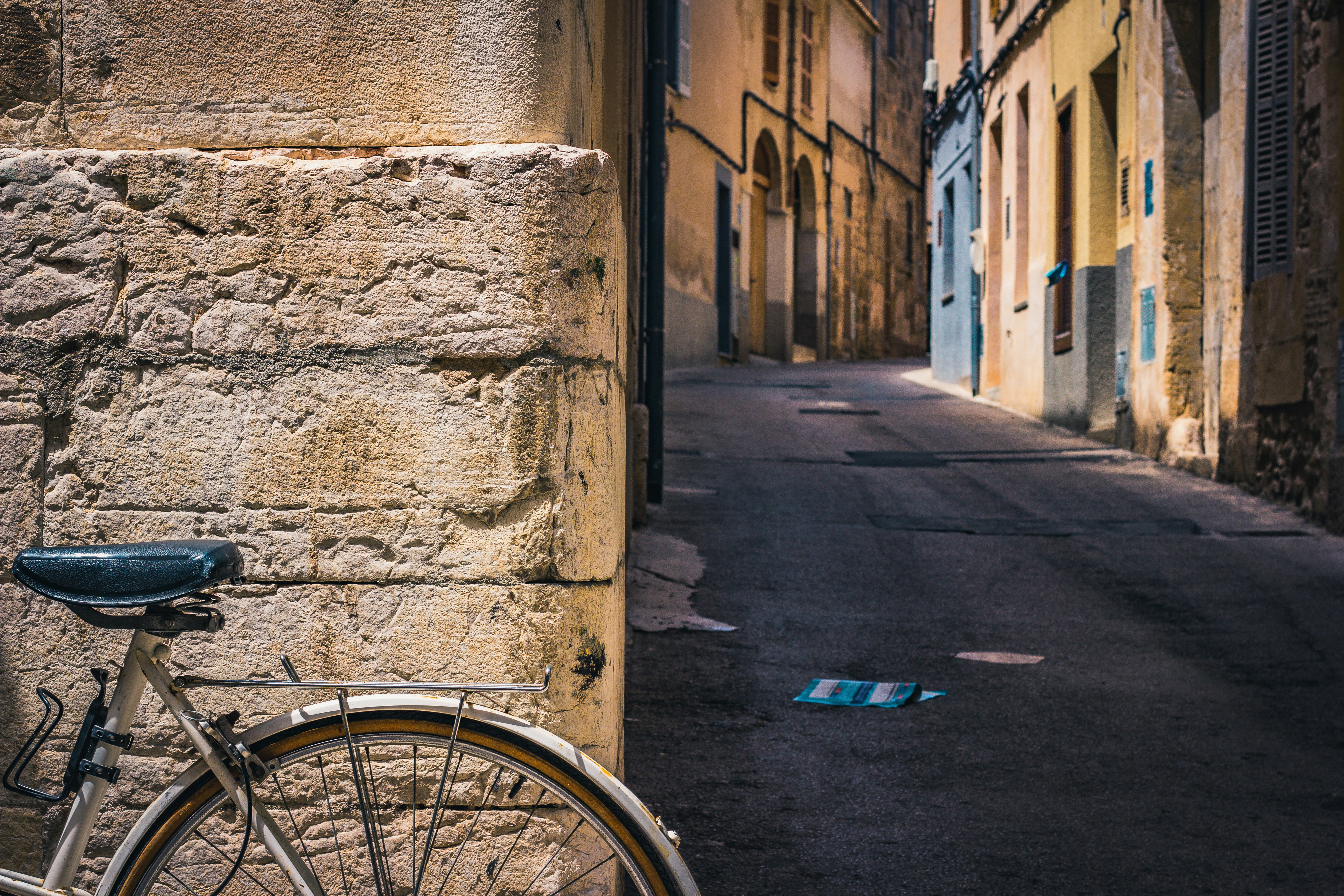 A vintage bicycle leans against a weathered stone wall in a sunlit alleyway, leading into a charming street adorned with pastel-colored buildings.