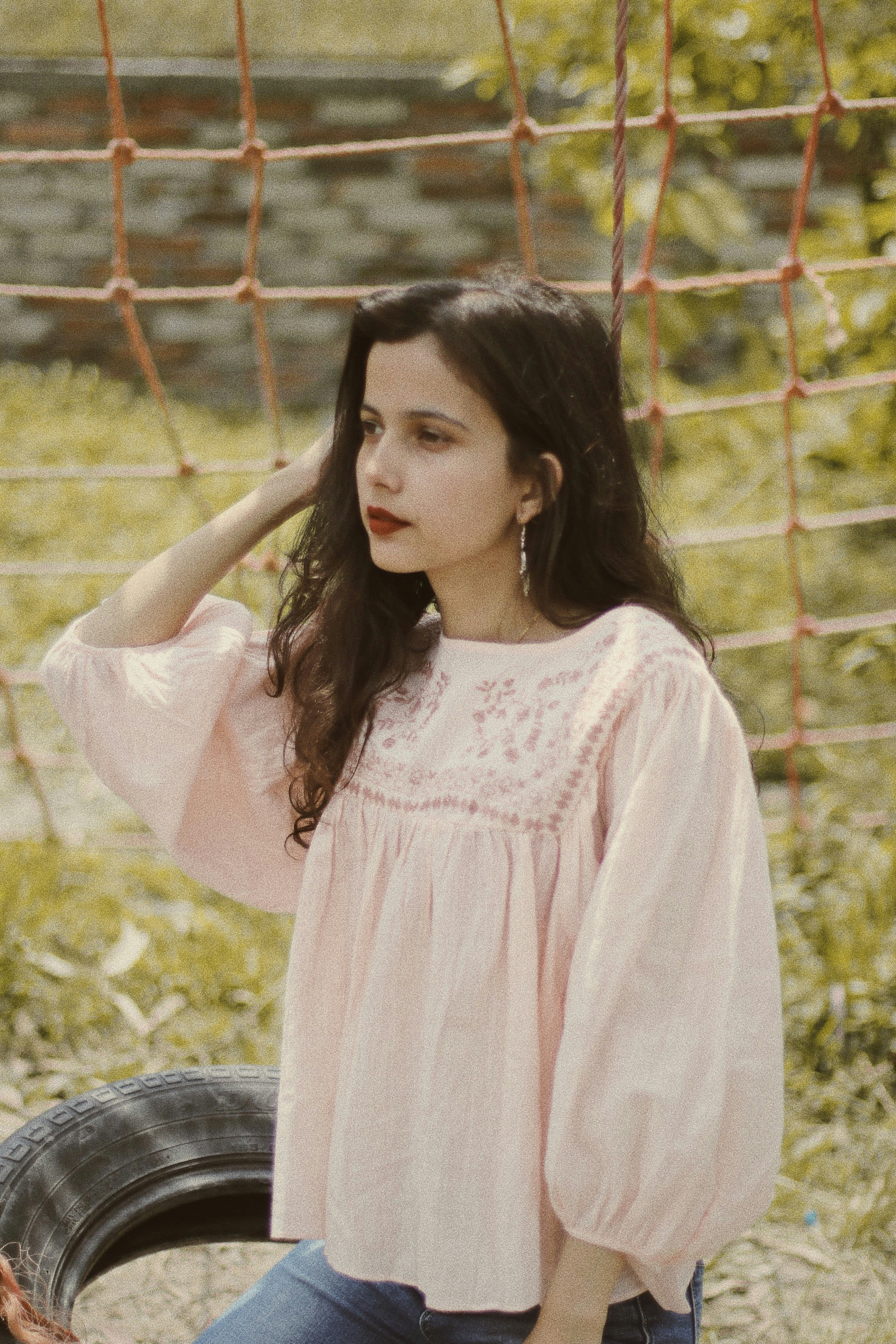 A young woman in a pastel pink blouse poses thoughtfully against a rustic backdrop, evoking a sense of nostalgia. The scene captures a moment of serene reflection amidst nature.