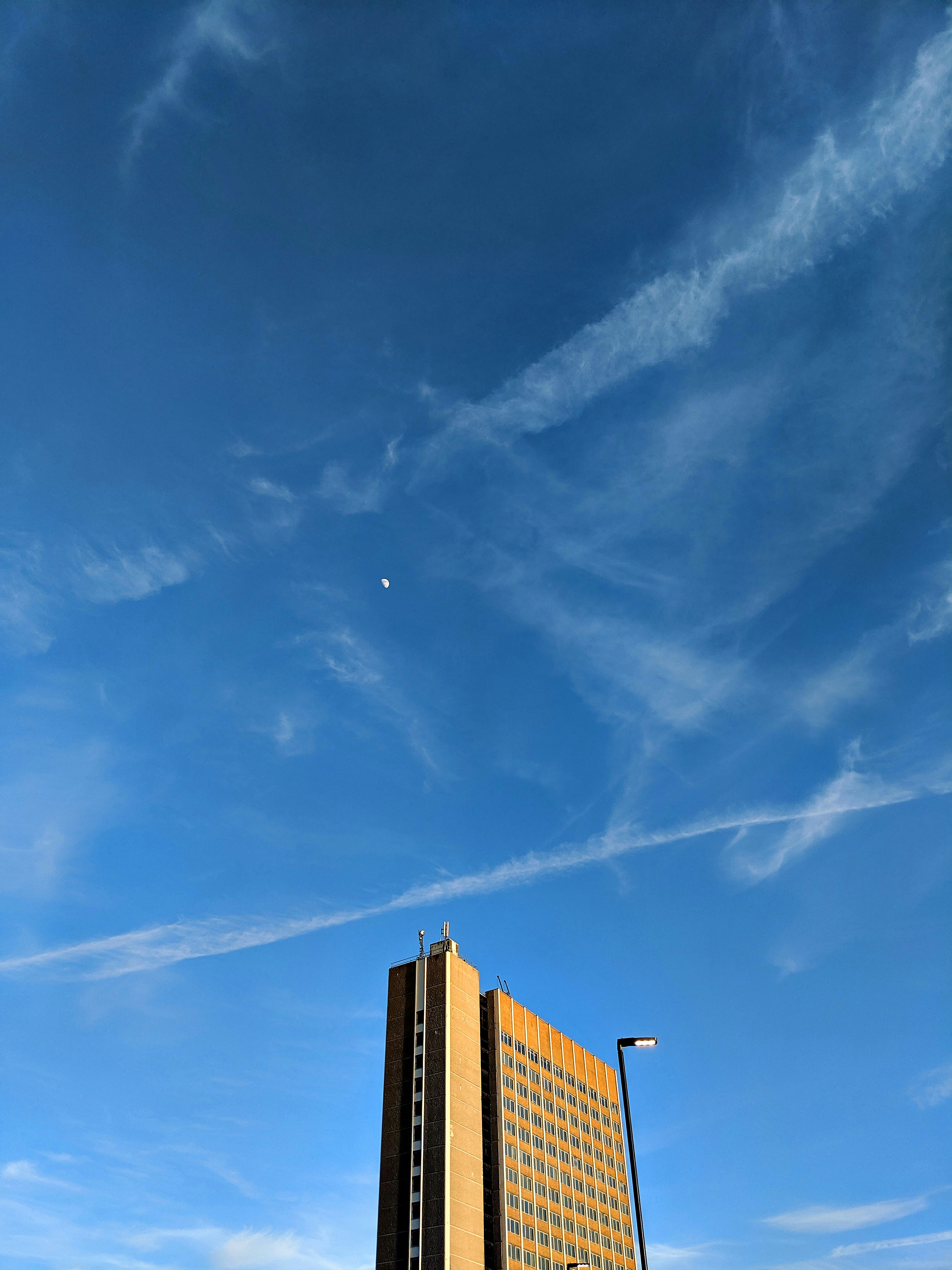 A tall building rises against a vibrant blue sky adorned with wispy clouds, while a small moon hangs above, adding a touch of serenity.