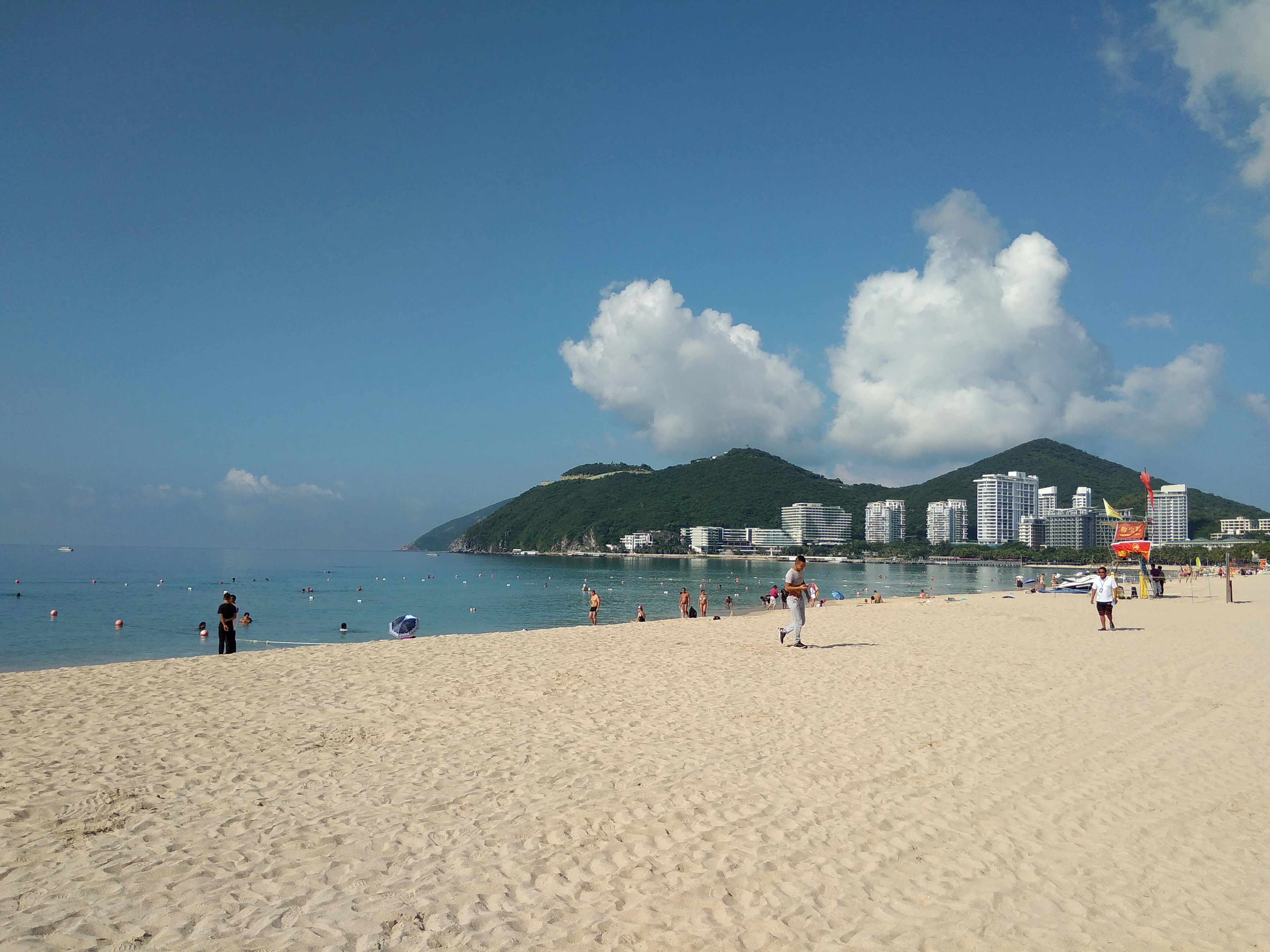 Vibrant beach scene with sunbathers and swimmers enjoying the clear waters, framed by distant mountains and modern buildings.