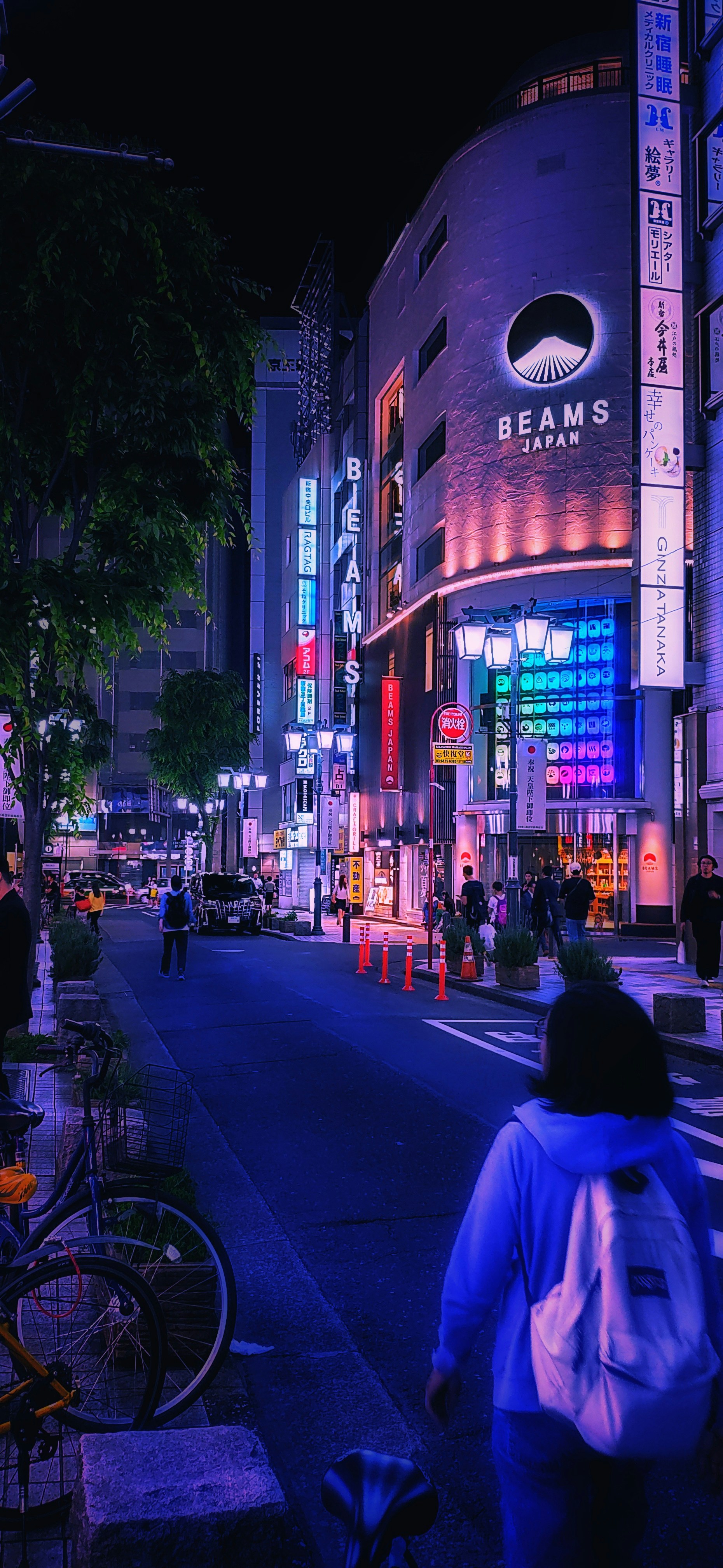 Nighttime street scene in Tokyo with neon signs and the Beams Japan building glowing purple and blue. Pedestrians and bicycles line the curb as a passerby with a backpack walks away from the camera.
