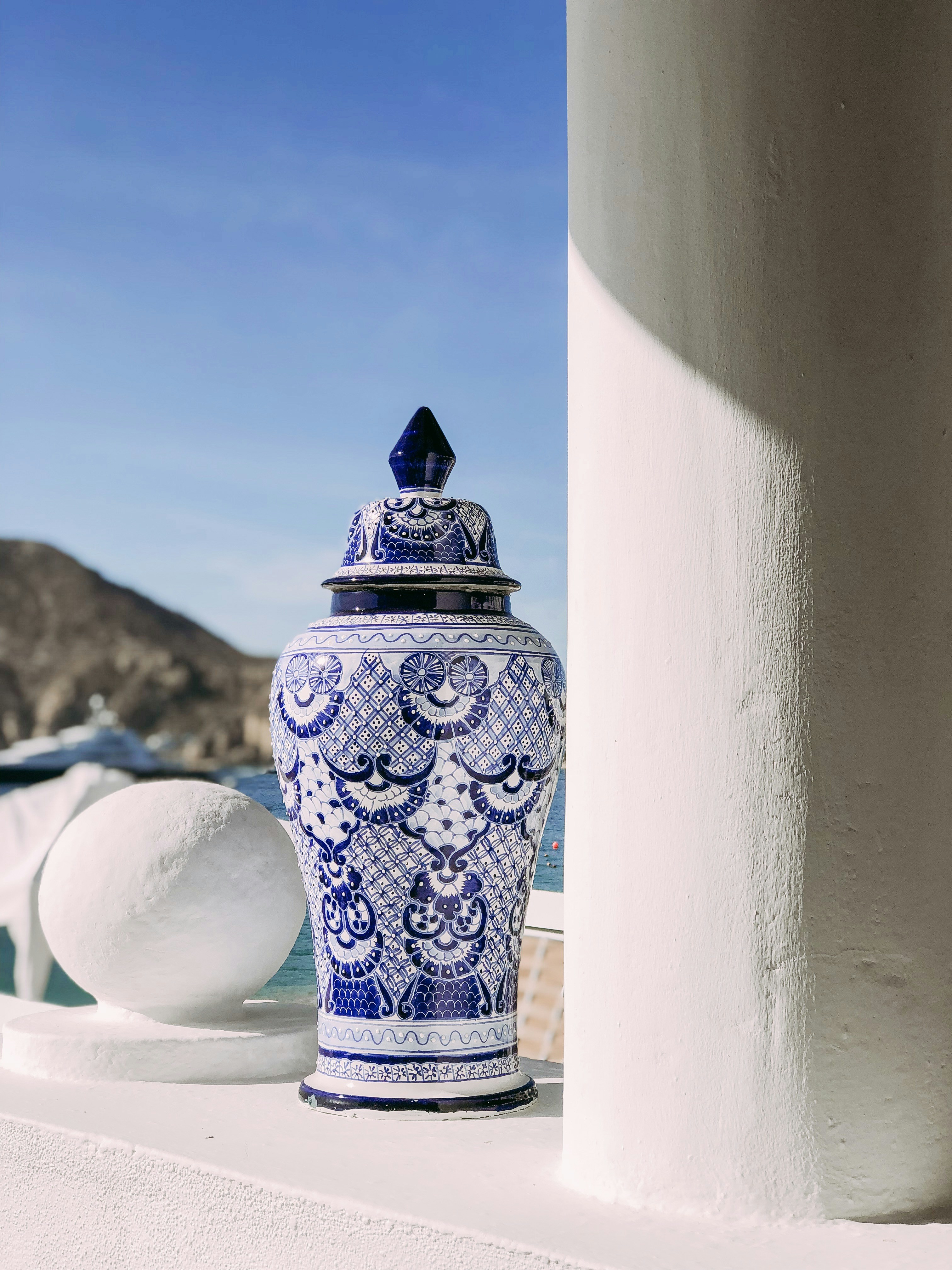 Intricately designed blue and white ceramic urn beside a smooth white column, with a coastal backdrop under a clear sky.