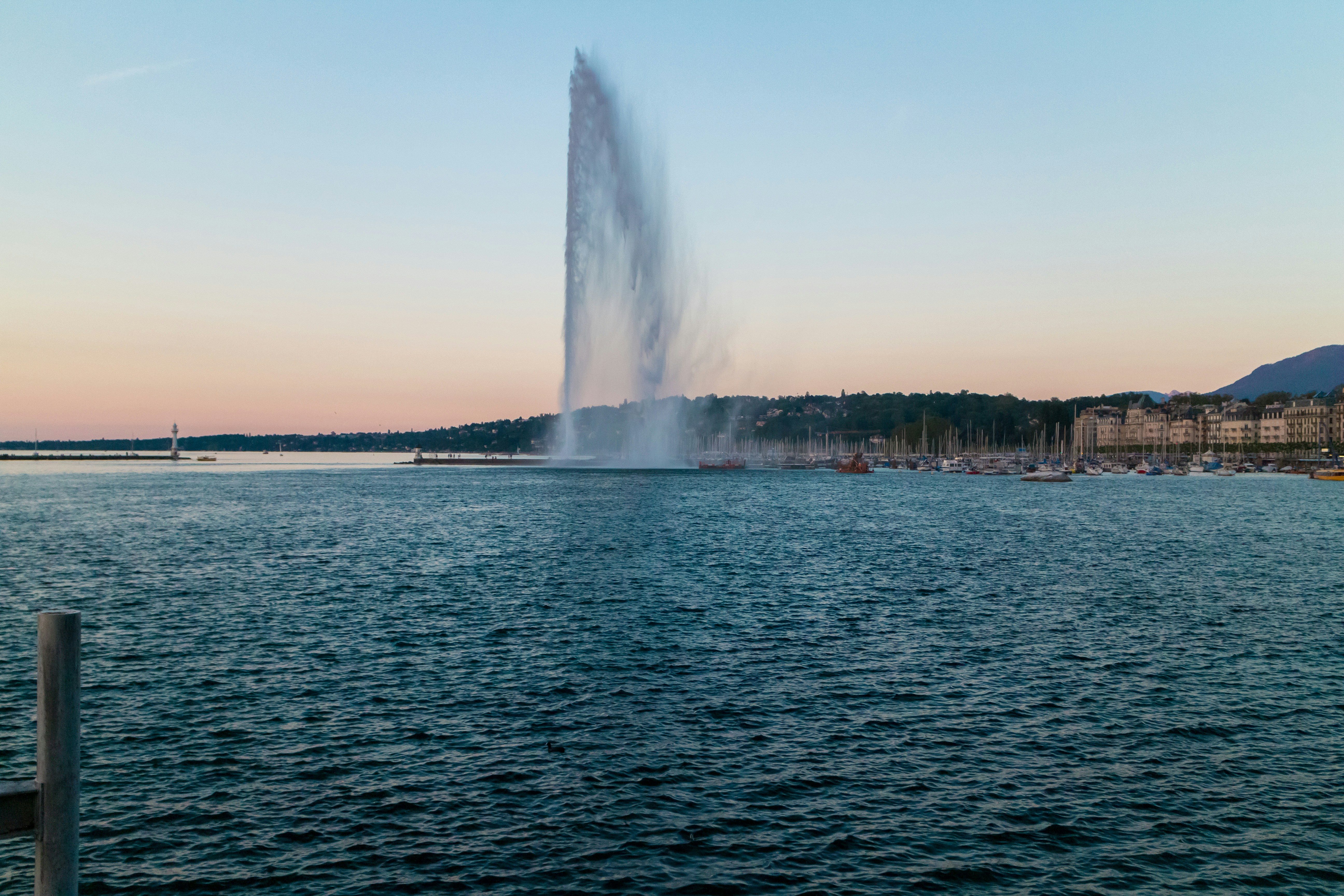 Dusk Jet Over the Harbor: A Tall Fountain Pierces the Calm Water Against a Sunset Skyline