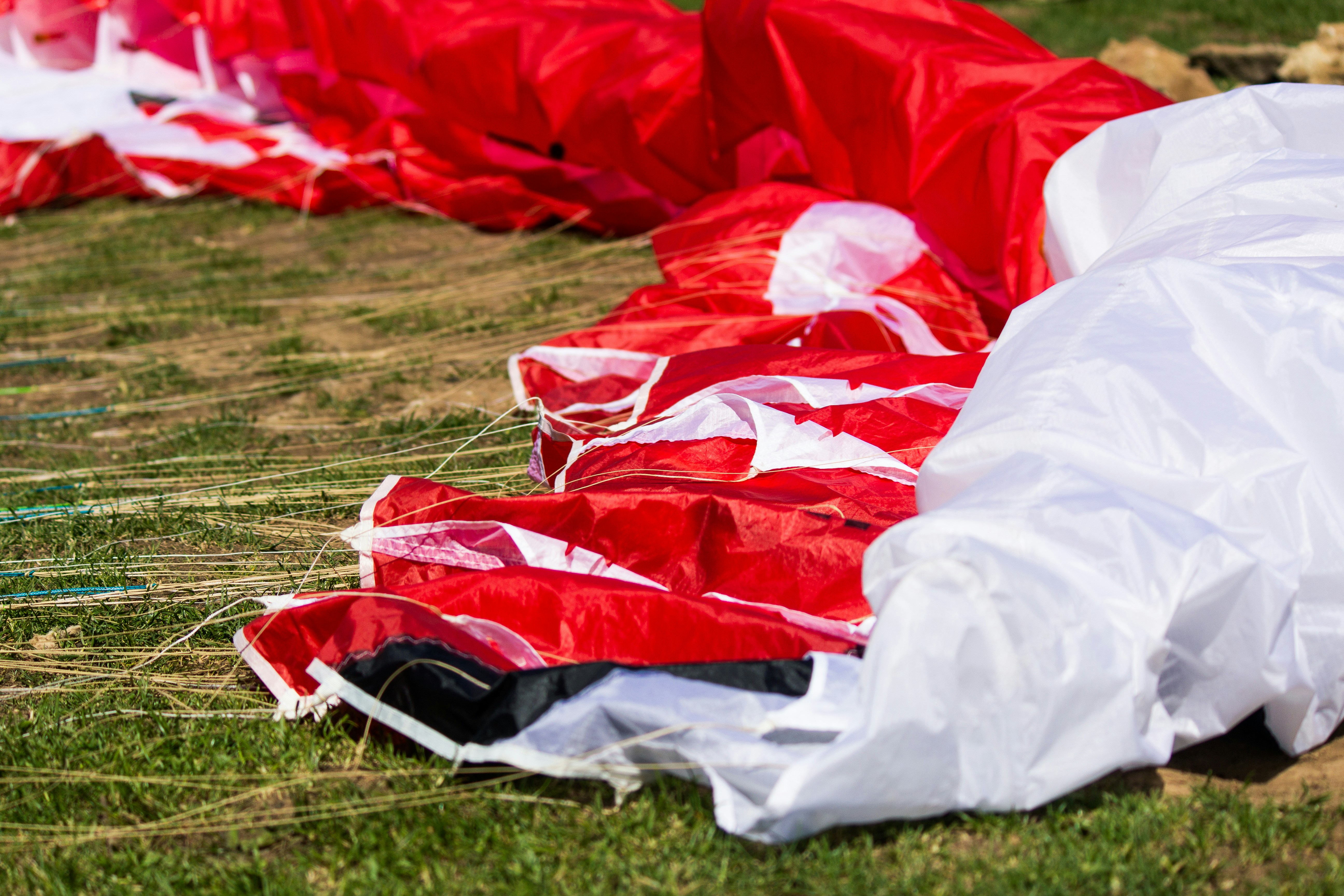Colorful paragliding canopies, red and white, laid out on green grass, ready for flight. The intricate lines and folds showcase the preparation for an aerial adventure.