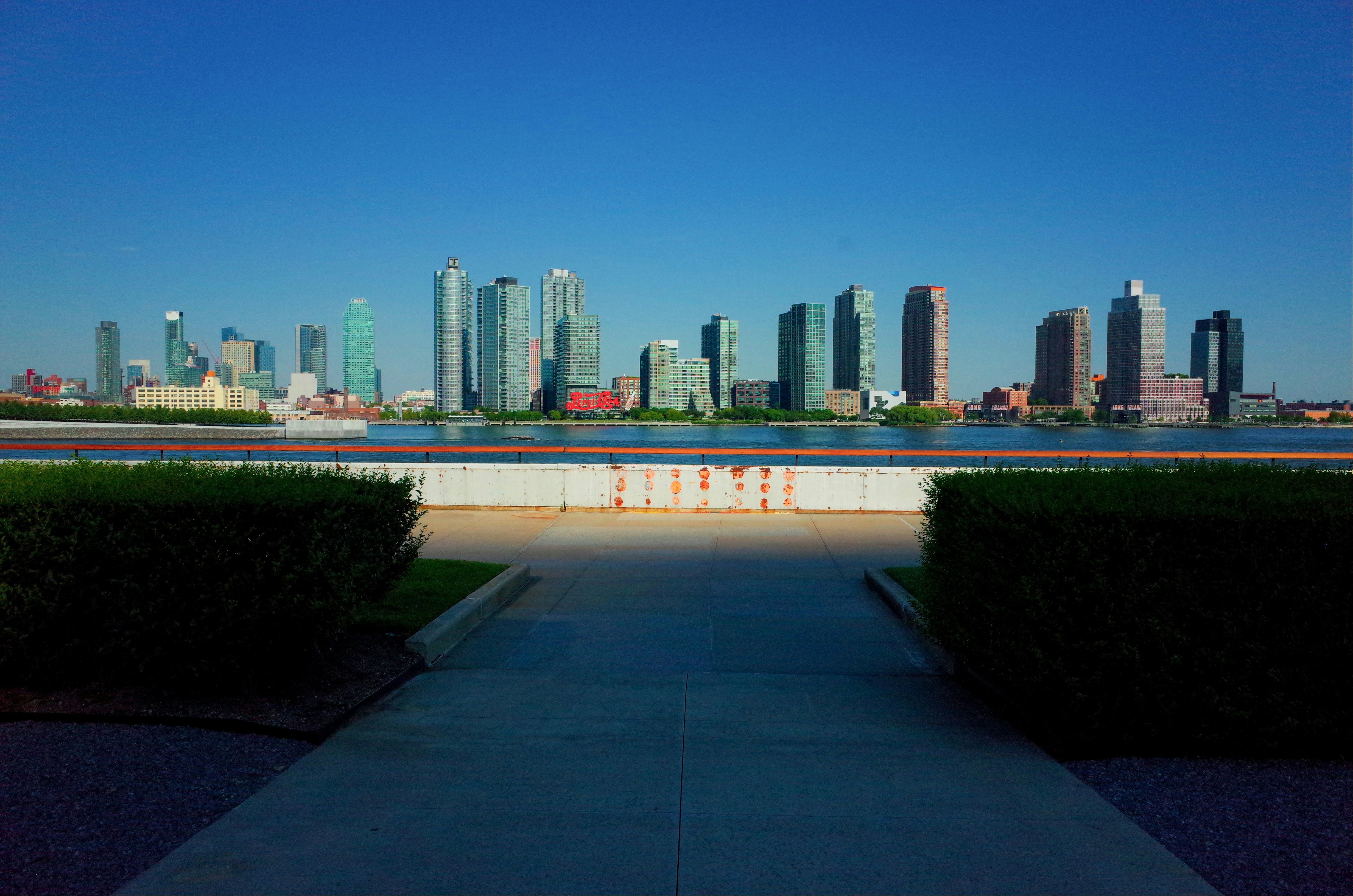 A vibrant city skyline reflects in the water, framed by lush greenery in the foreground. The scene captures the essence of urban life against a clear blue sky.