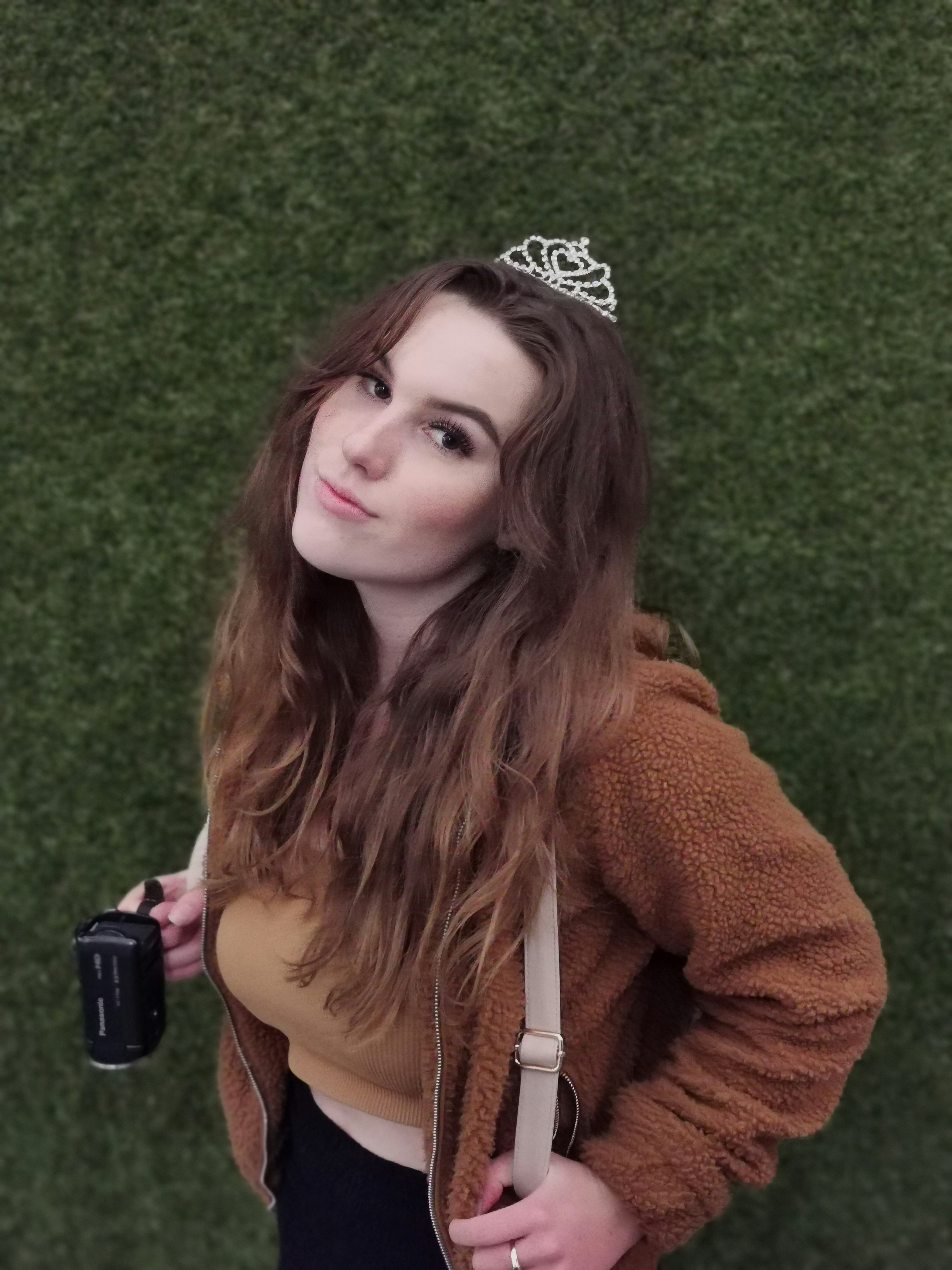 Woman in a brown textured jacket and tiara stands against a lush green wall, holding a camera.