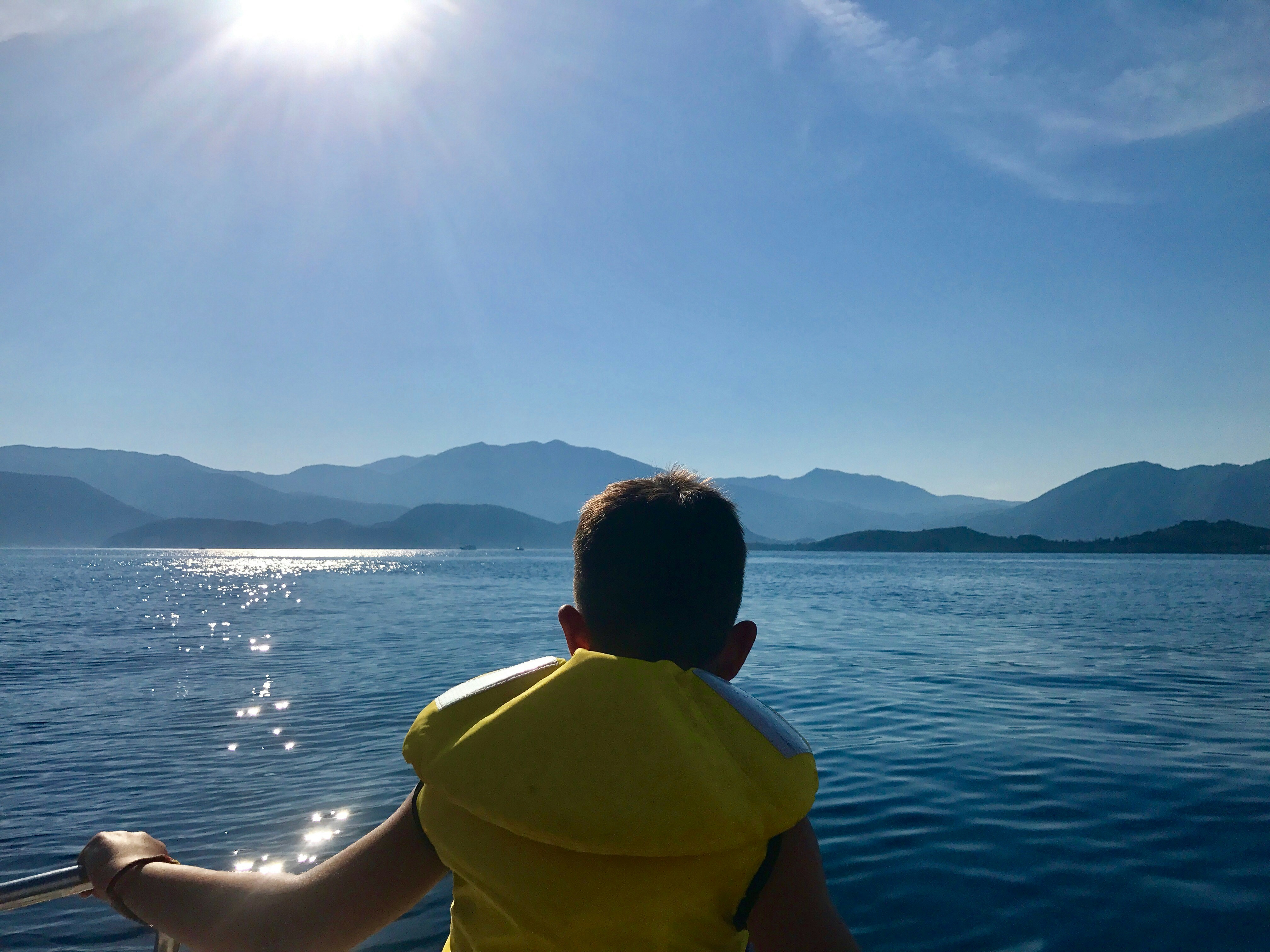 Child in a yellow life jacket gazing at the tranquil sea and distant mountains under a bright sun.