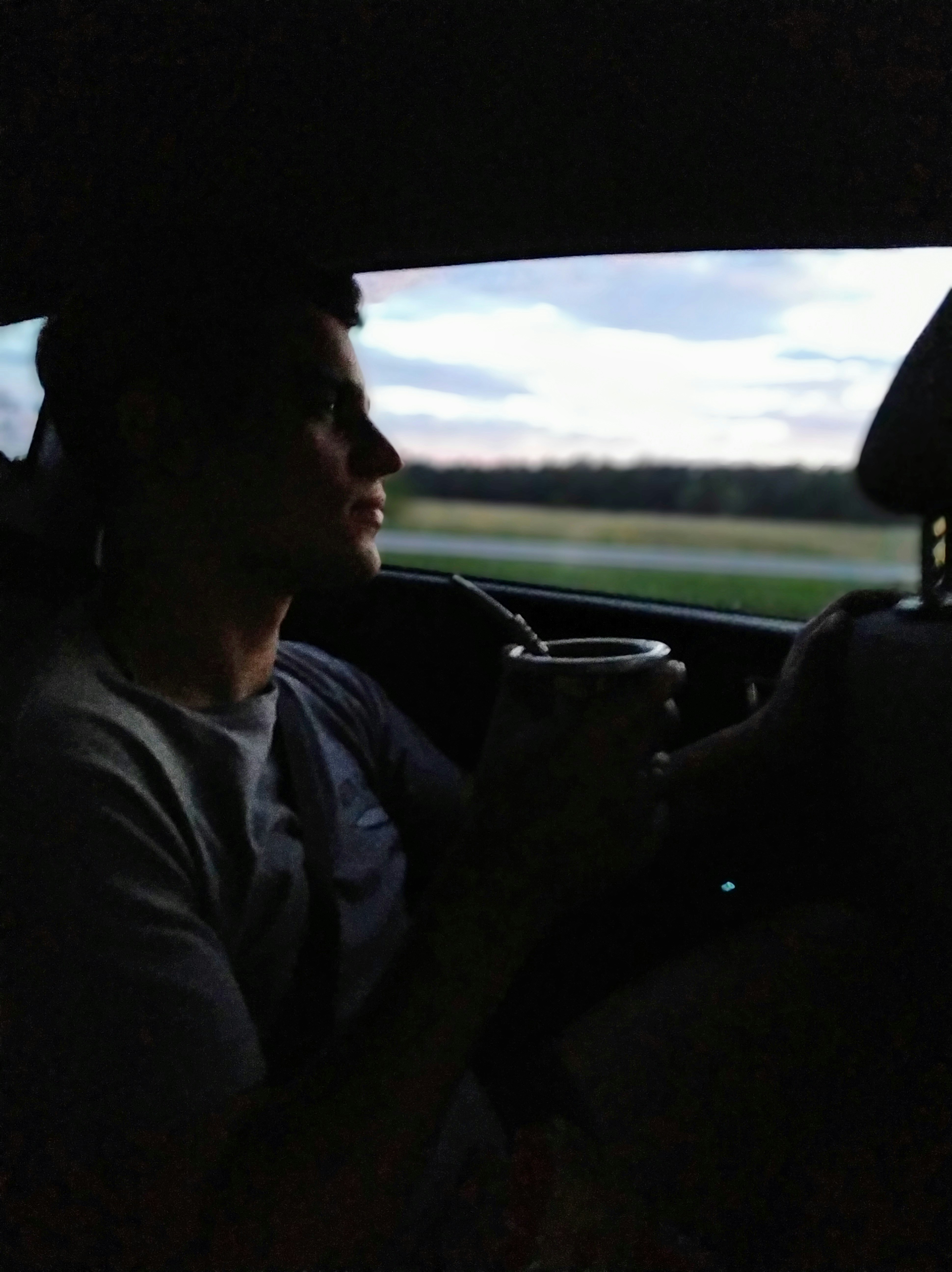 Dimly lit photograph of a man in a car, holding a cup, with a blurred landscape visible through the window.
