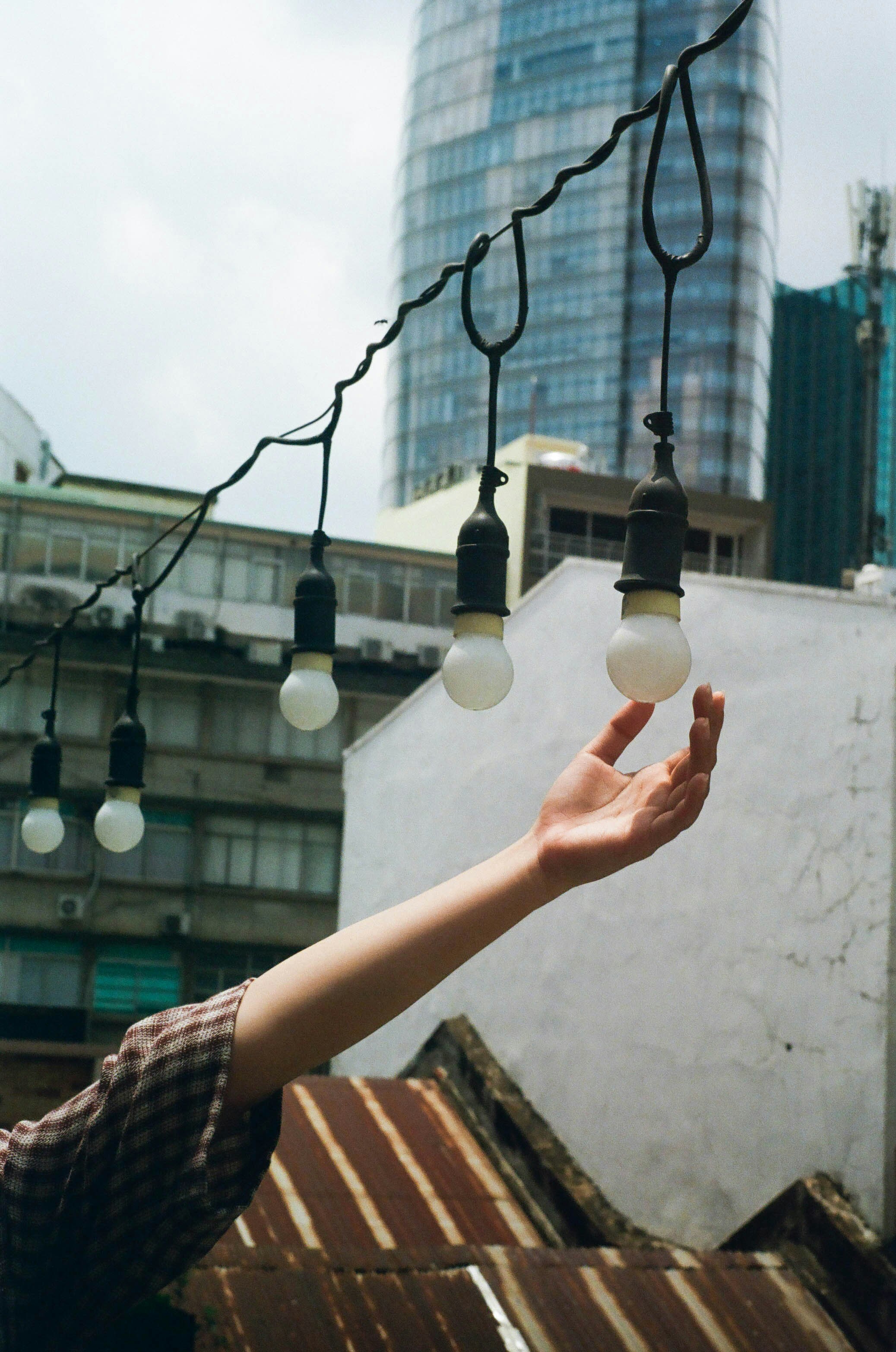 A hand reaches towards hanging light bulbs against a backdrop of modern architecture and weathered rooftops.