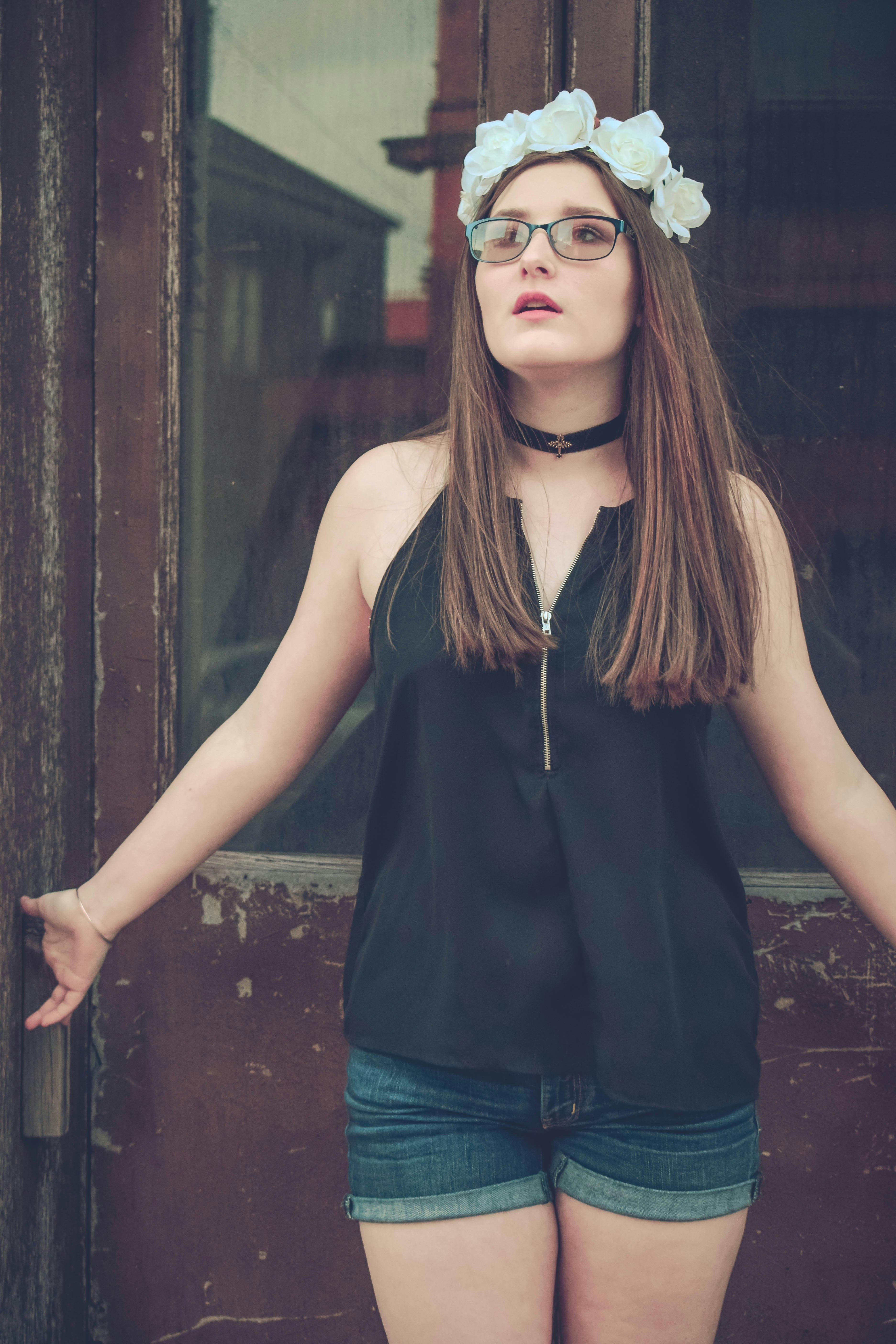 A young woman stands confidently in front of a weathered door, adorned with a floral crown and stylish attire, embodying a blend of modernity and nature.