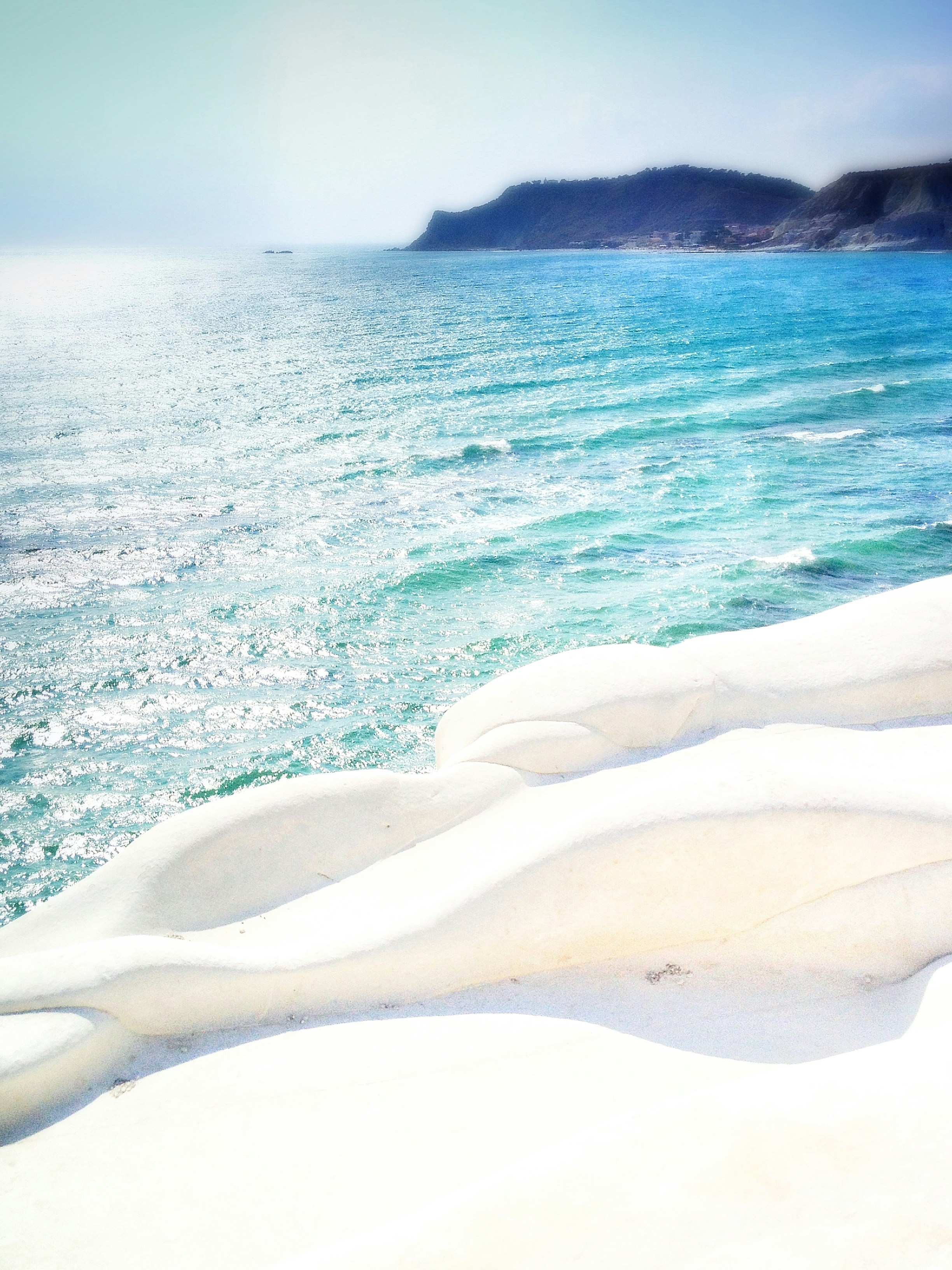 Bright coastal photograph featuring smooth white rock formations in the foreground, turquoise sea stretching toward a distant headland under clear light.