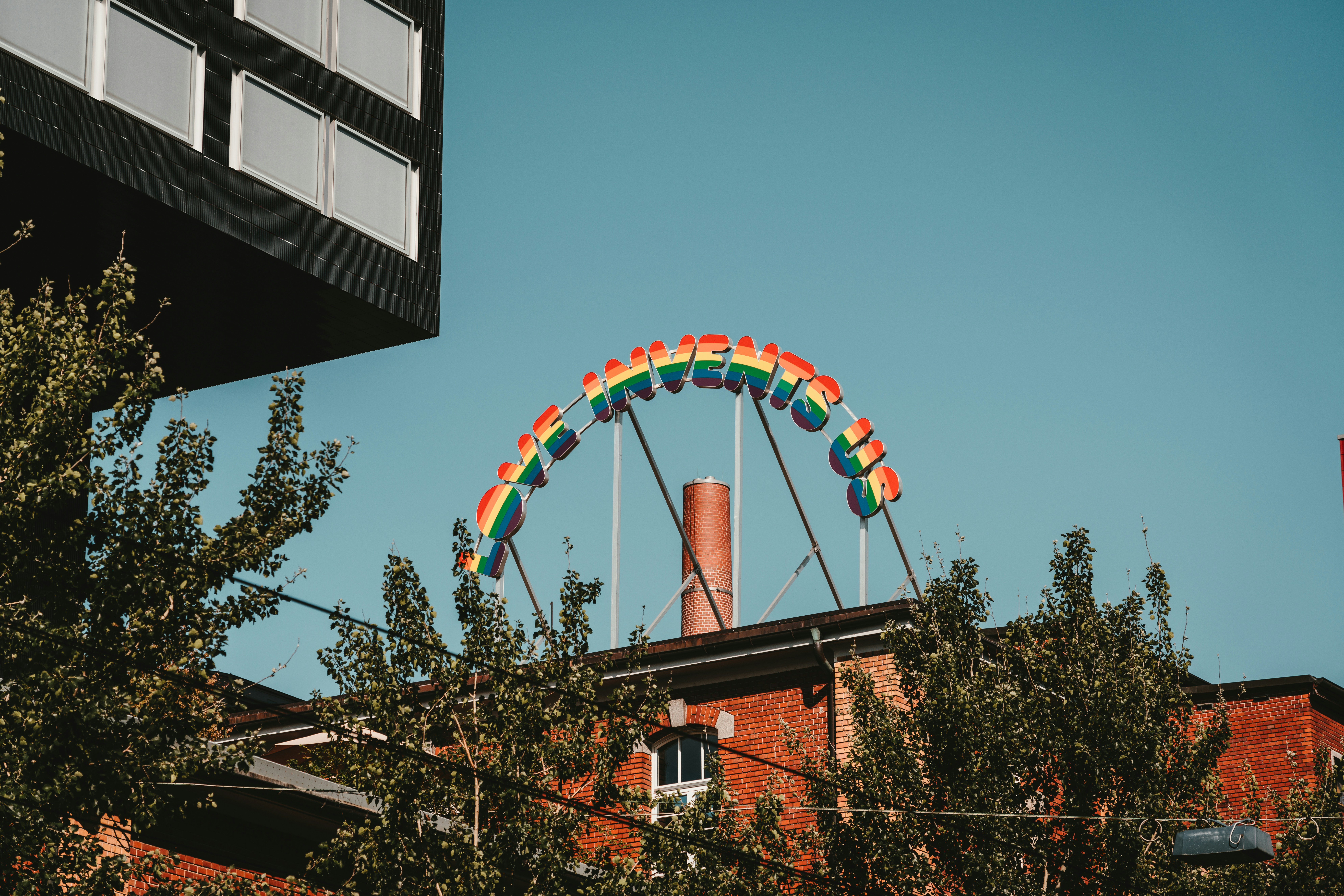 Colorful rainbow arch sign atop a brick building, framed by urban foliage and modern architecture.