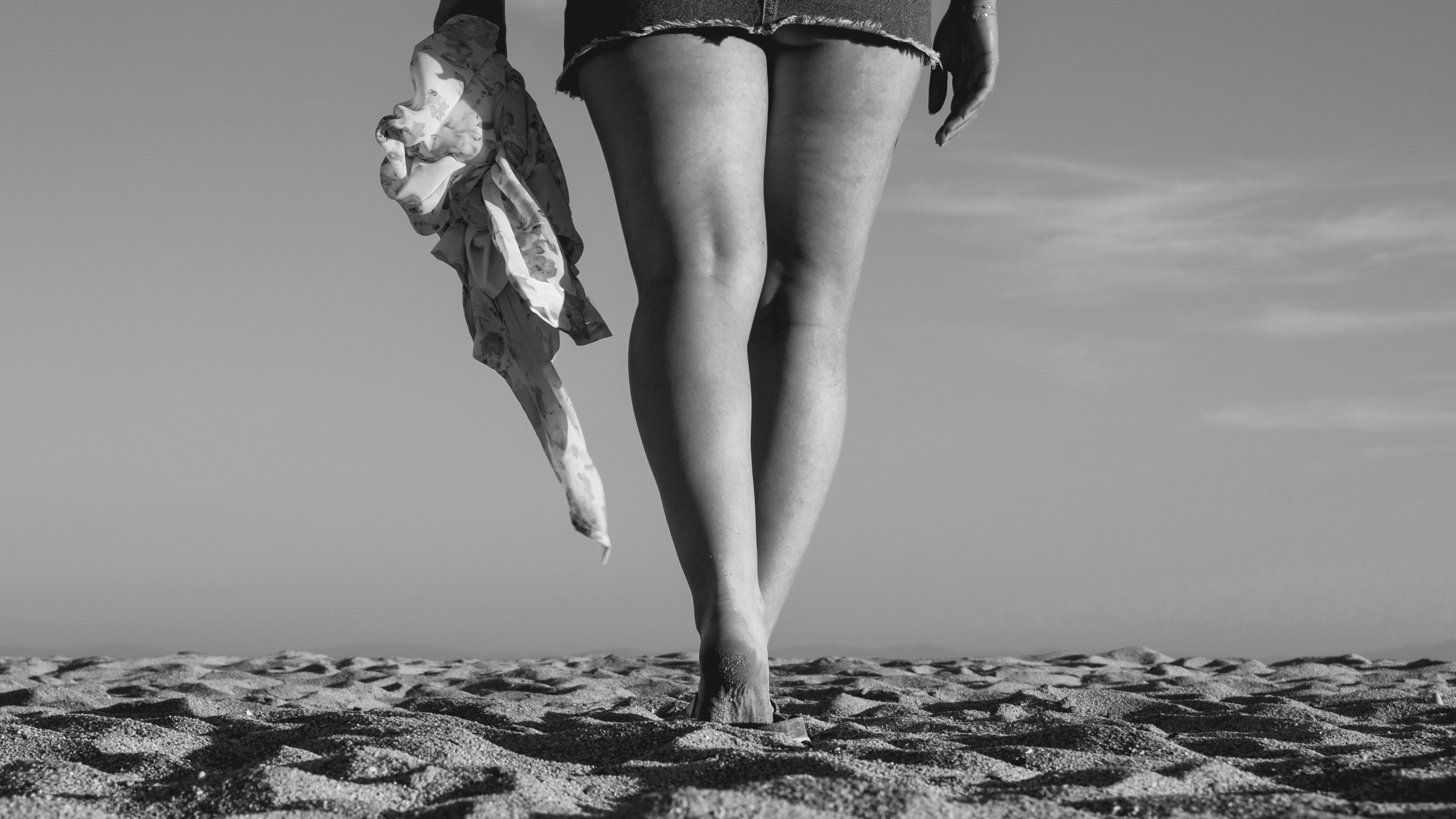 A figure walks barefoot on the sandy beach, holding a light garment, captured in black and white to emphasize texture and form.