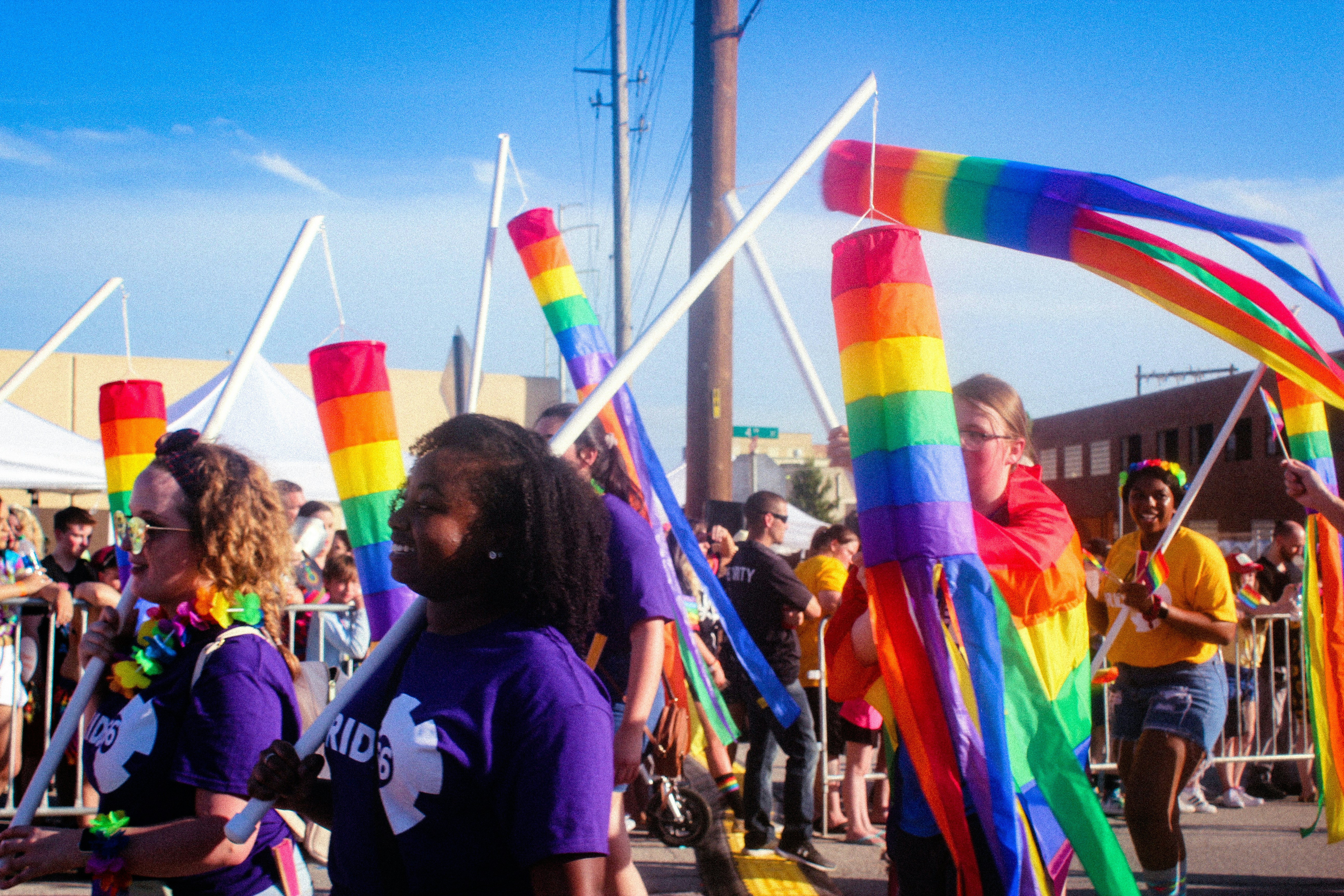 Group of people marching with rainbow flags in a lively street parade under a clear blue sky.