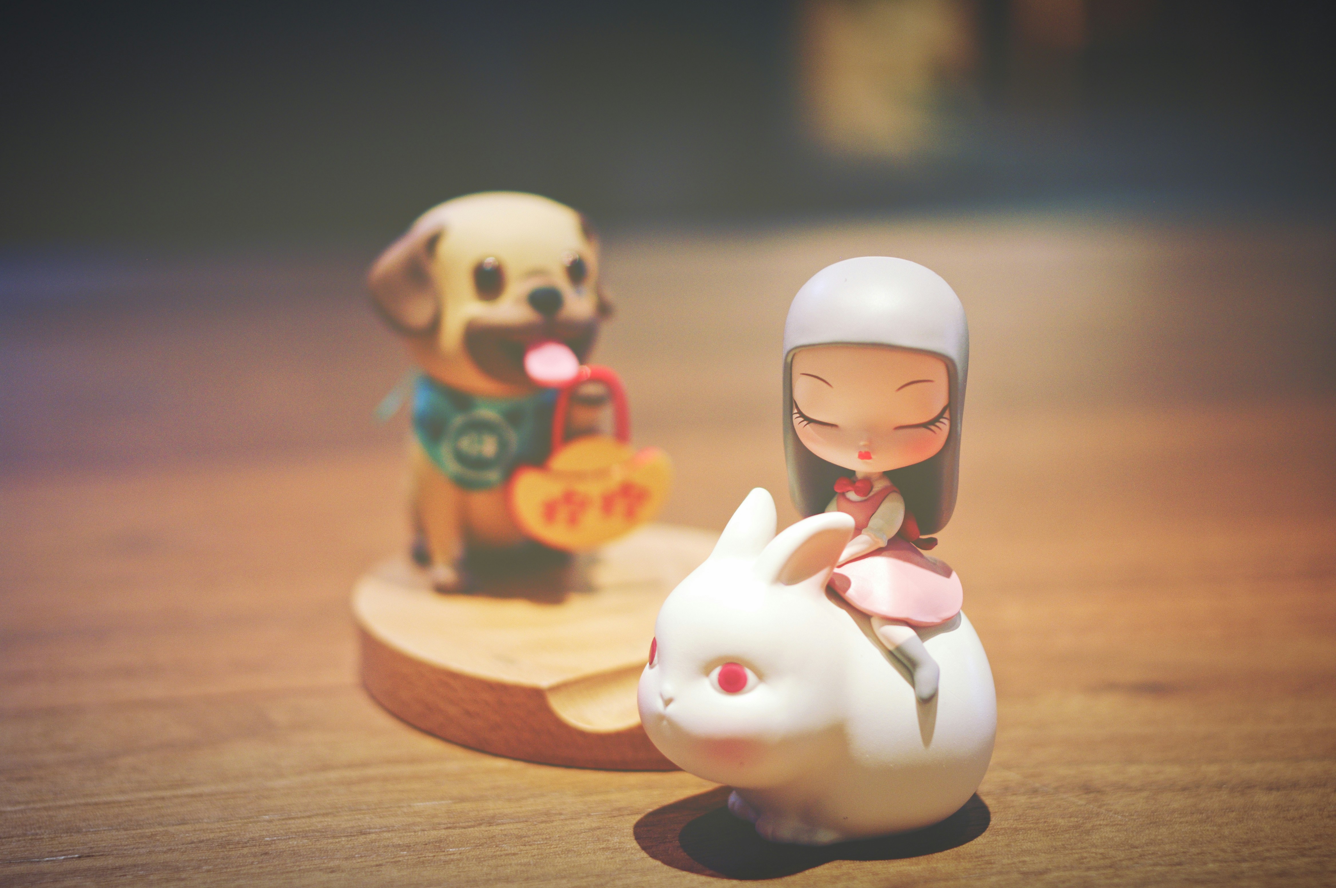 Photograph of a whimsical miniature scene featuring a girl riding a white rabbit toy, with a small dog figure in the blurred background. The shallow depth of field isolates the foreground, highlighting the charming setup.