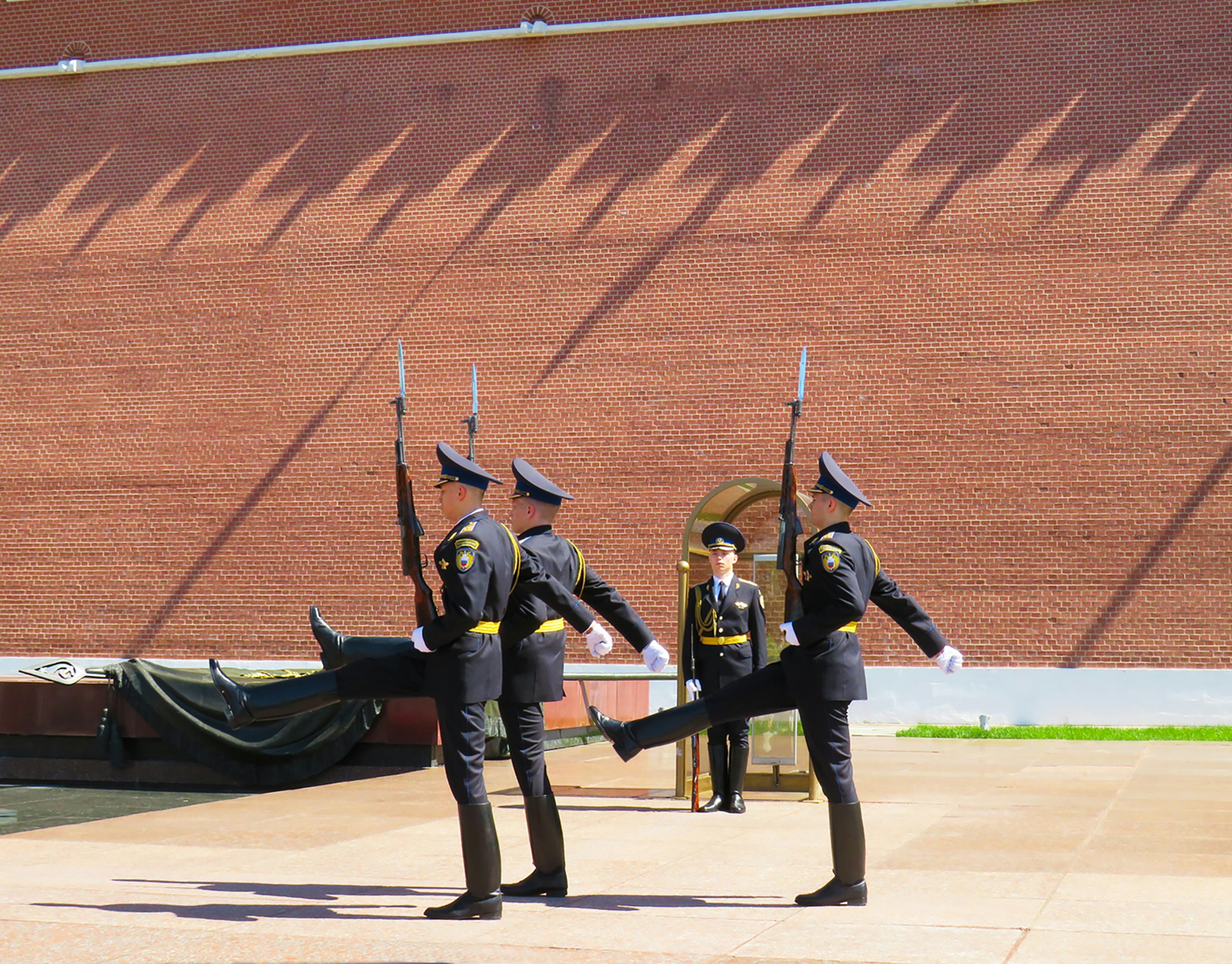 Soldiers in formal attire perform a synchronized drill with rifles in front of a historic brick wall, showcasing discipline and precision.