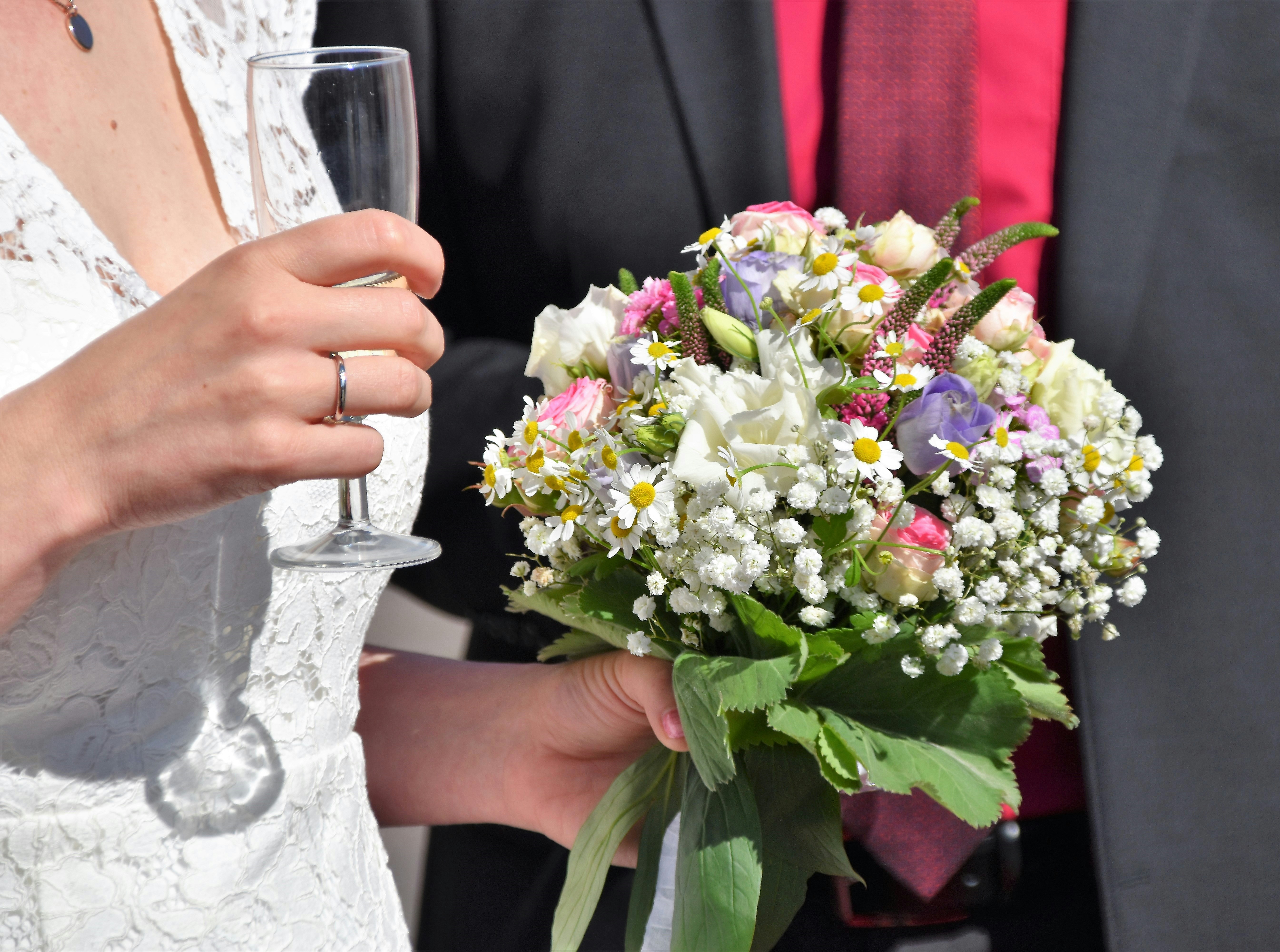 Bride holding a bouquet adorned with pastel flowers and greenery, while raising a champagne flute. A formal attire figure stands beside her.