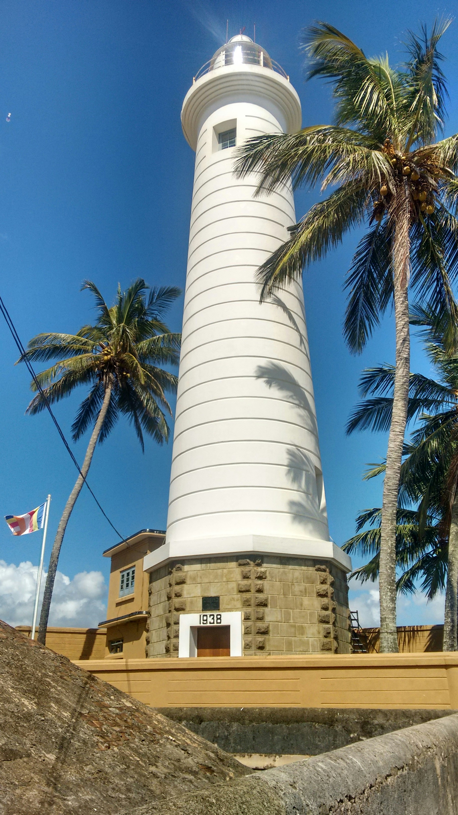 Historic lighthouse with a white tower and palm trees swaying in the breeze, set against a clear blue sky. The year 1938 is prominently displayed at its base.