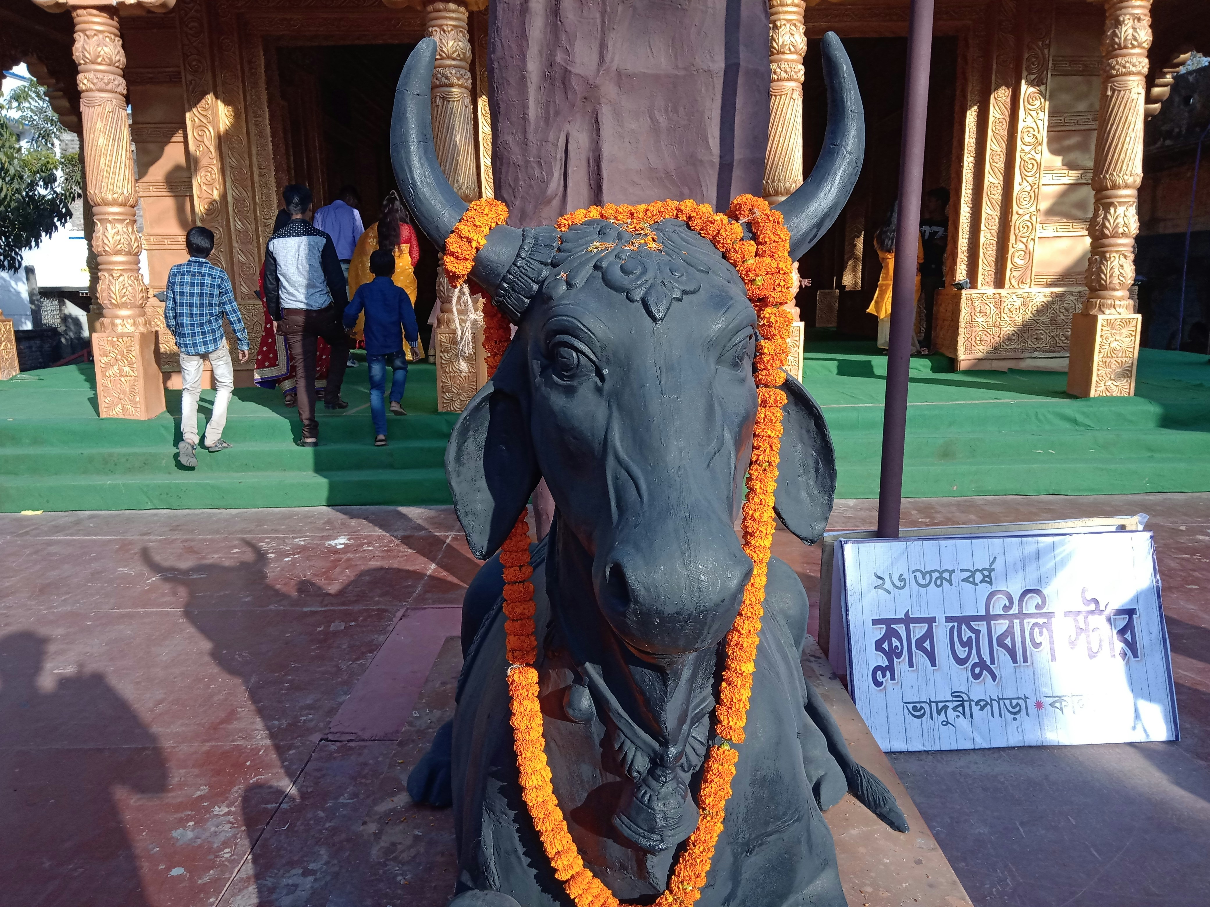Stone Nandi statue adorned with marigold garlands sits at temple entrance, with visitors in the background.