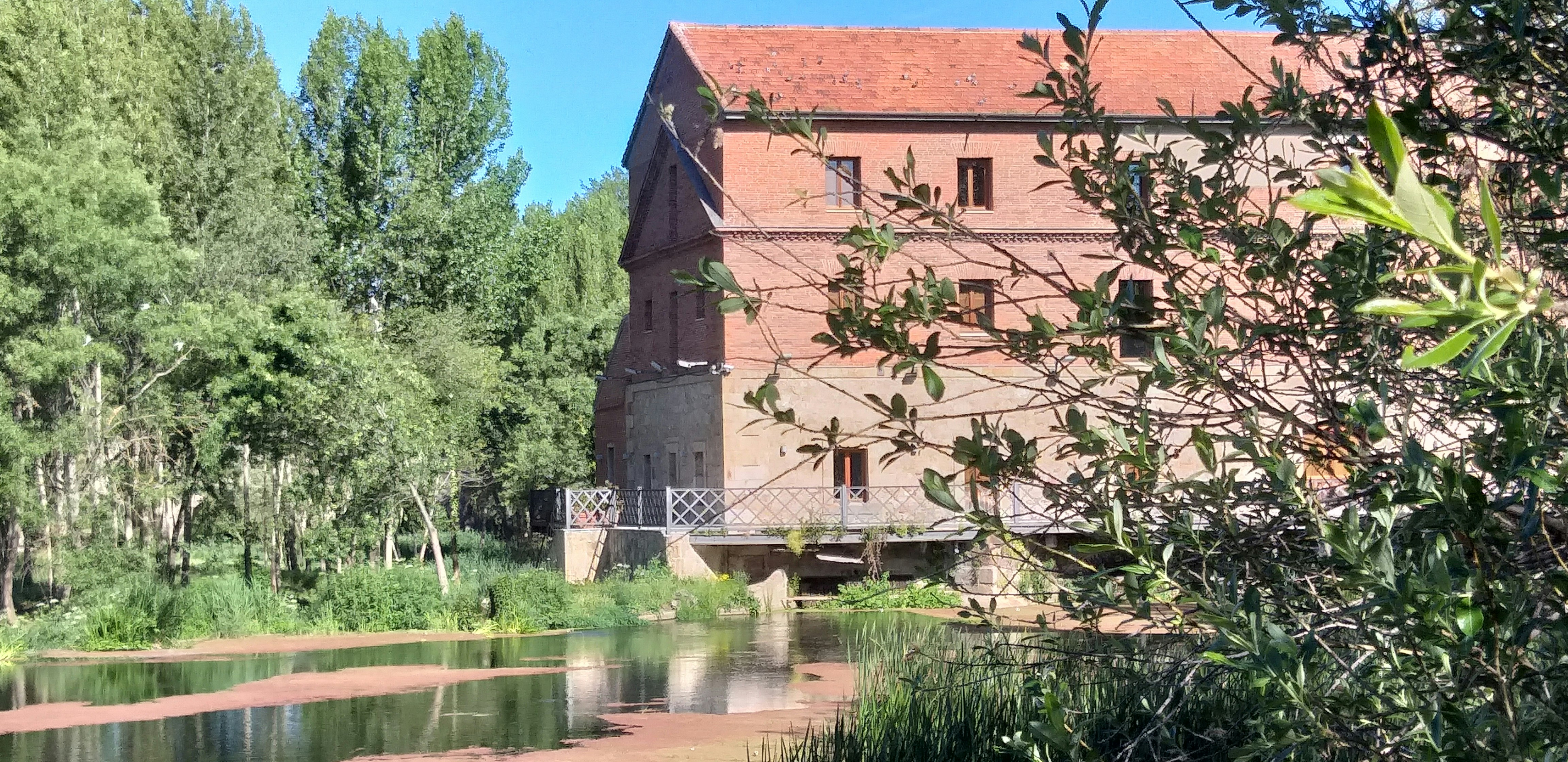 Historic brick building nestled beside a tranquil pond, surrounded by lush greenery and reflections on the water's surface.