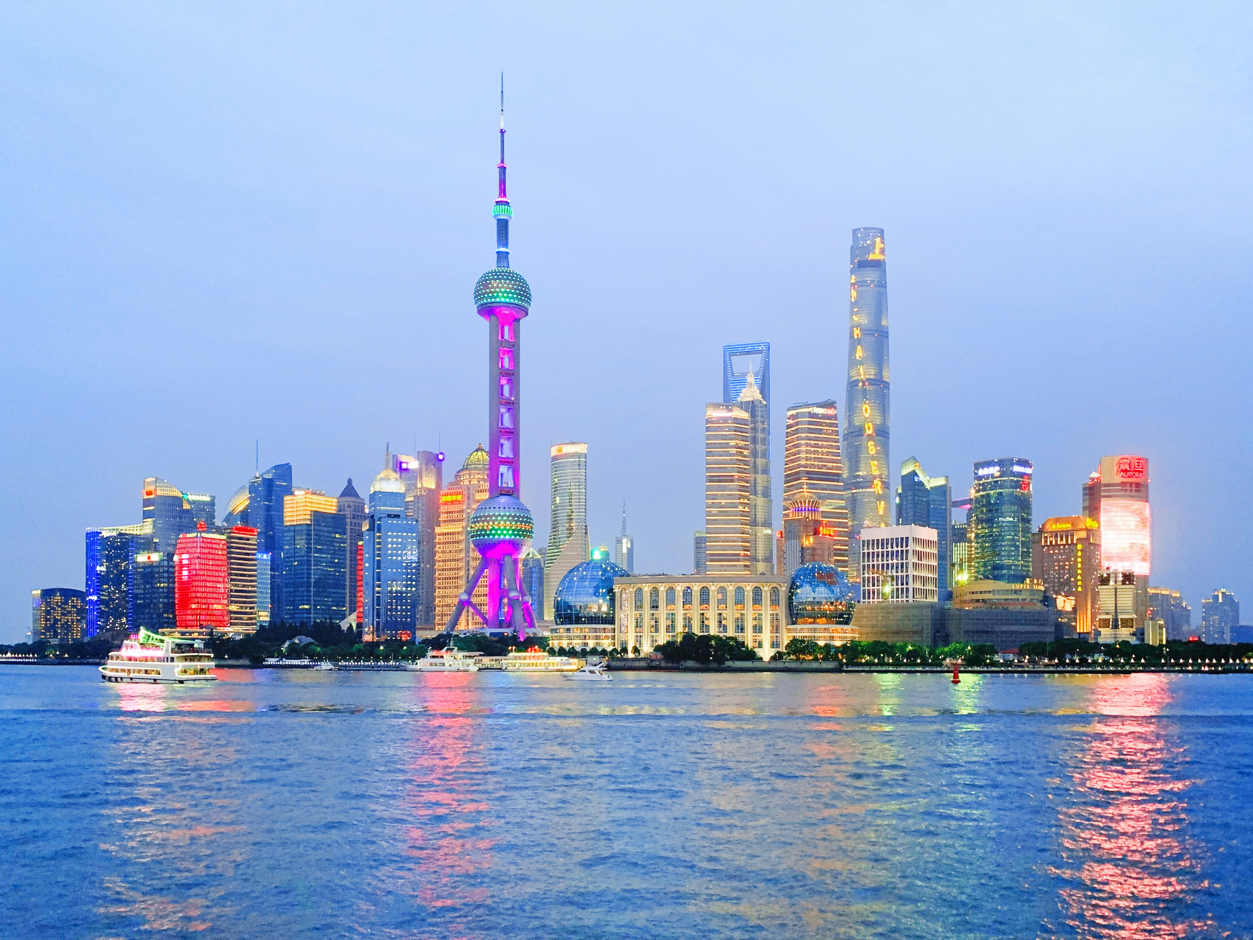 Vibrant skyline of Shanghai illuminated at dusk, showcasing modern architecture and the iconic Oriental Pearl Tower. A boat glides across the reflective waters.