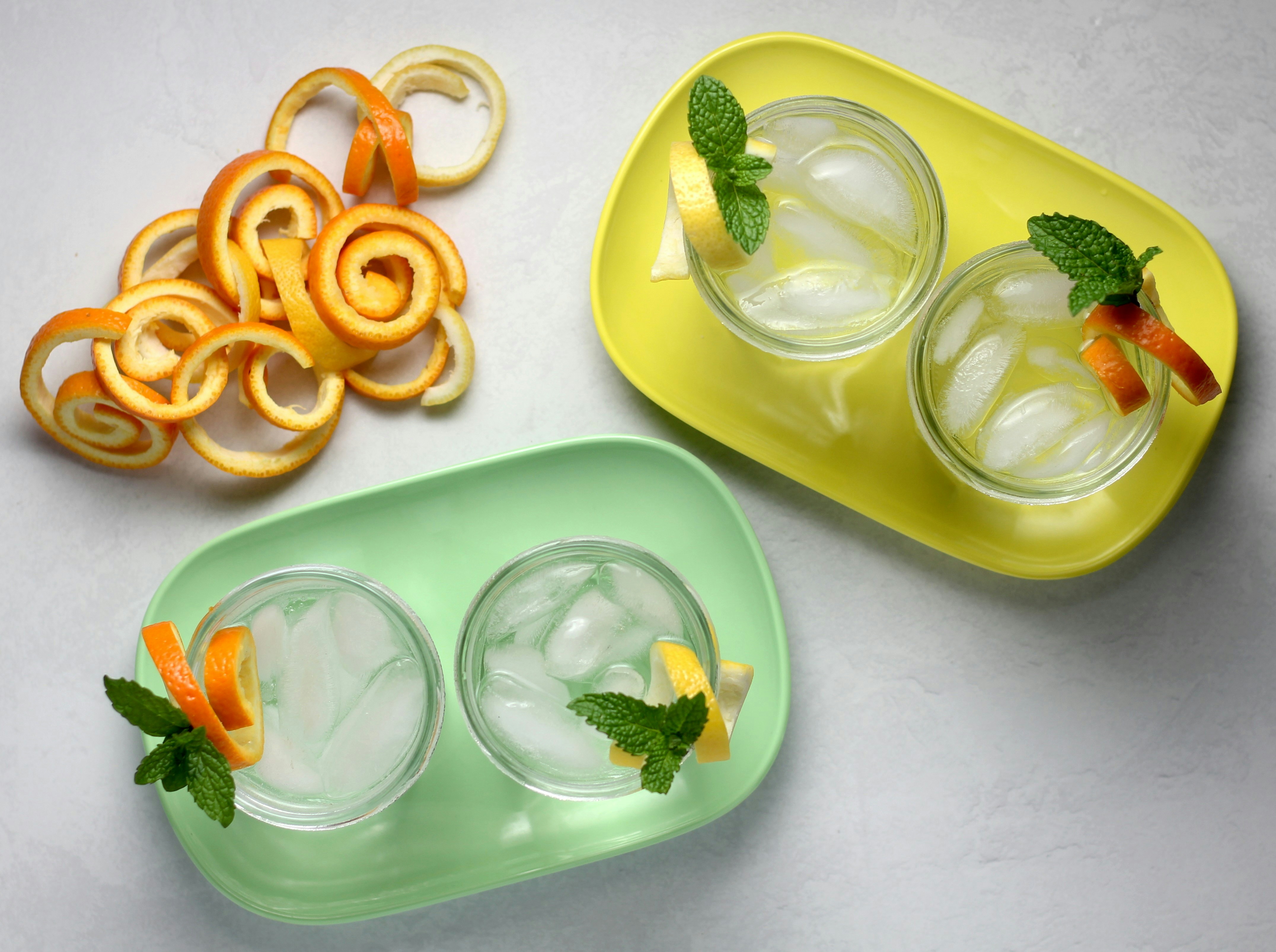 Two glasses filled with ice and garnished with citrus slices and mint leaves, accompanied by spirals of orange peel on a light surface.