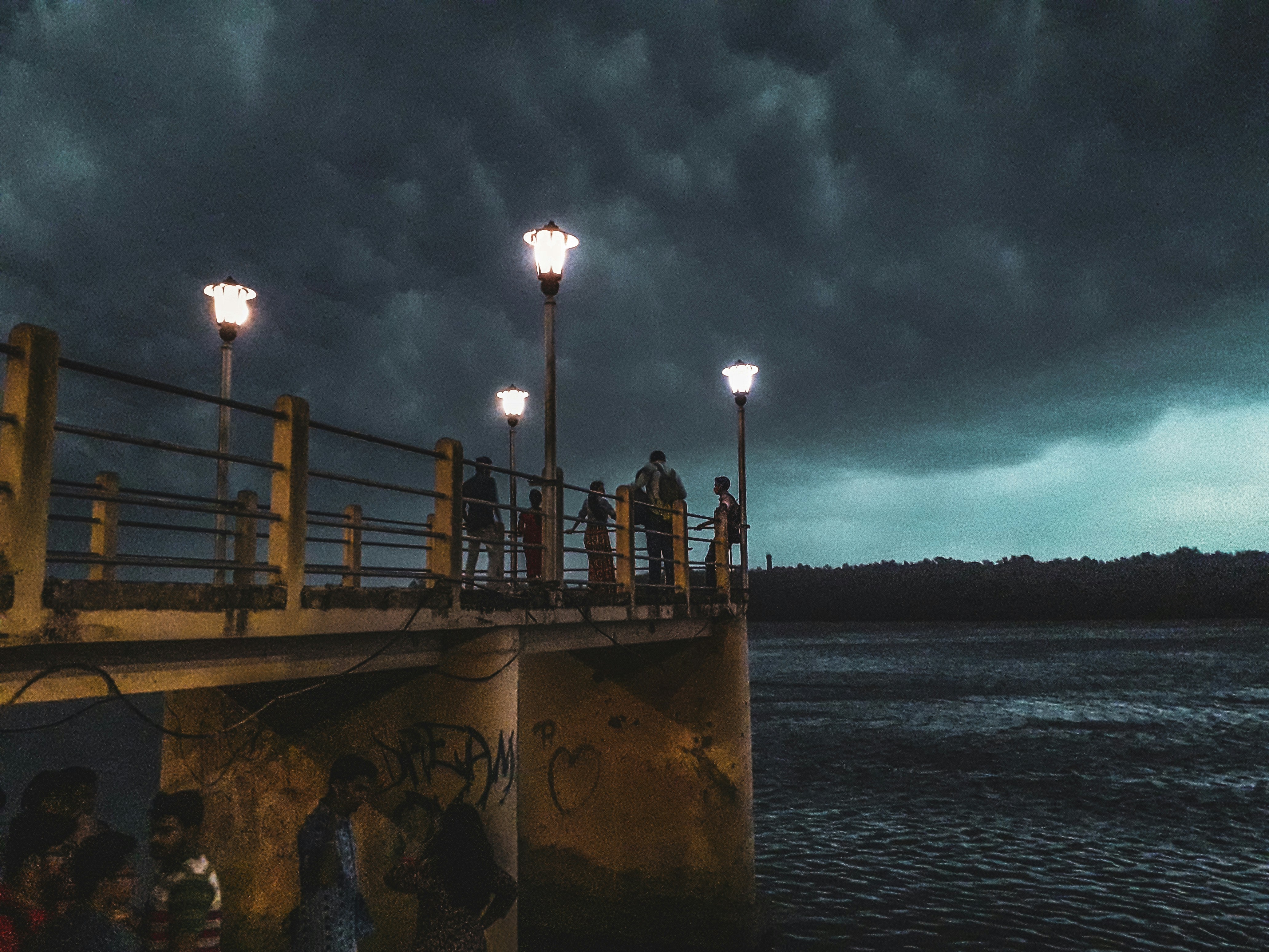 People standing on a pier illuminated by lampposts against a backdrop of dark storm clouds.