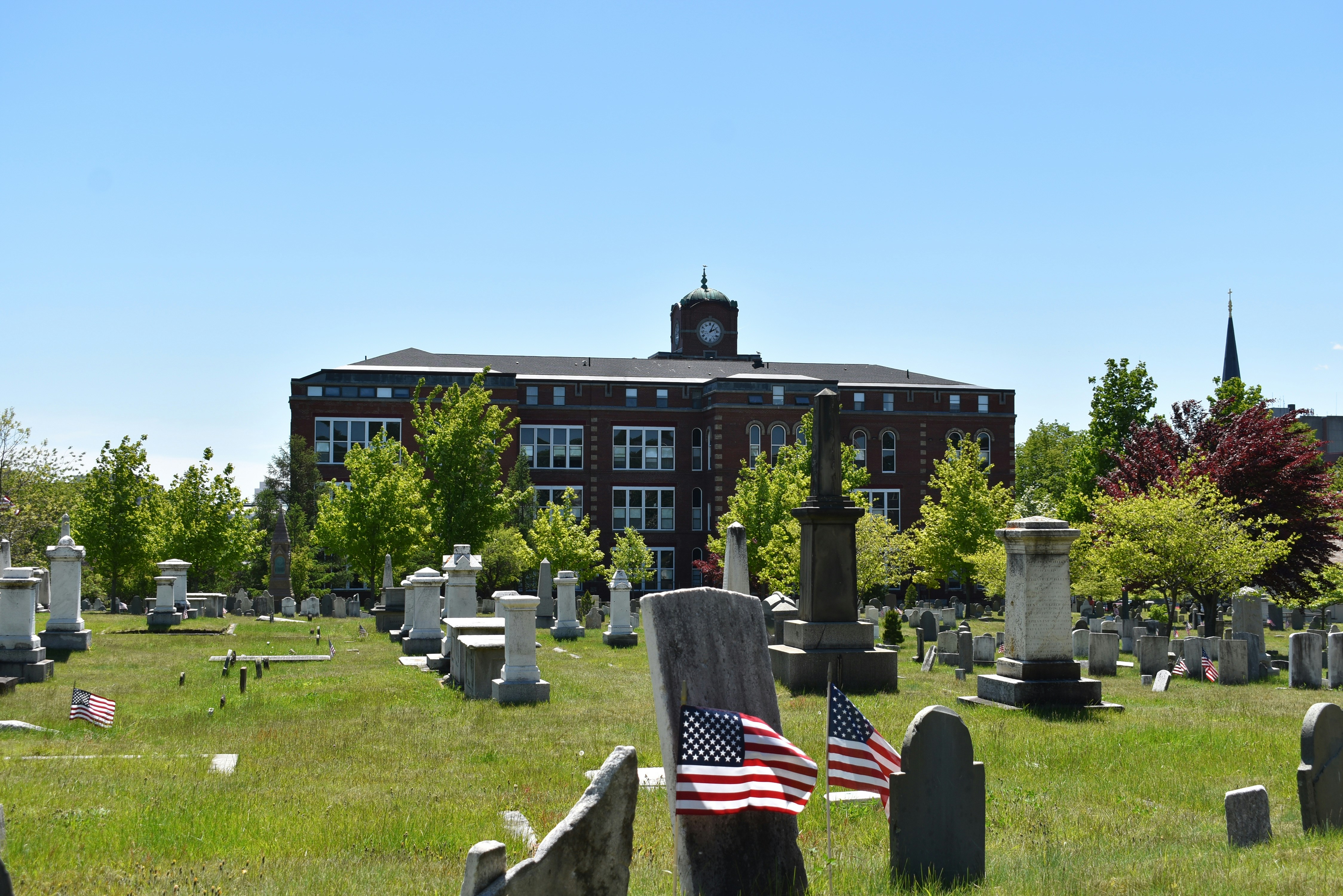 Gravestones adorned with American flags in a cemetery, framed by lush greenery and a historic building in the background.