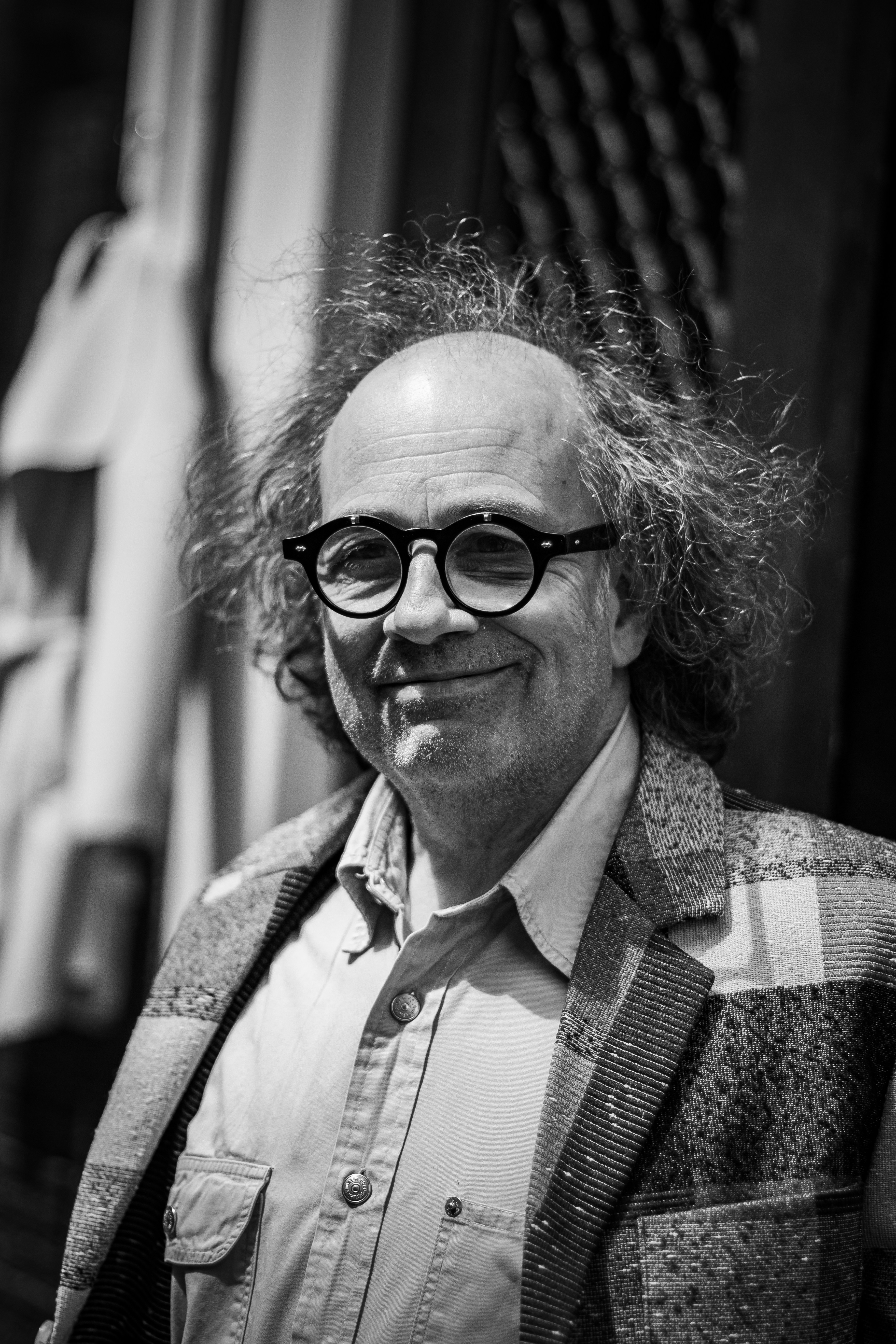 A middle-aged man with wild hair and glasses smiles warmly, dressed in a patterned blazer against a softly blurred background.