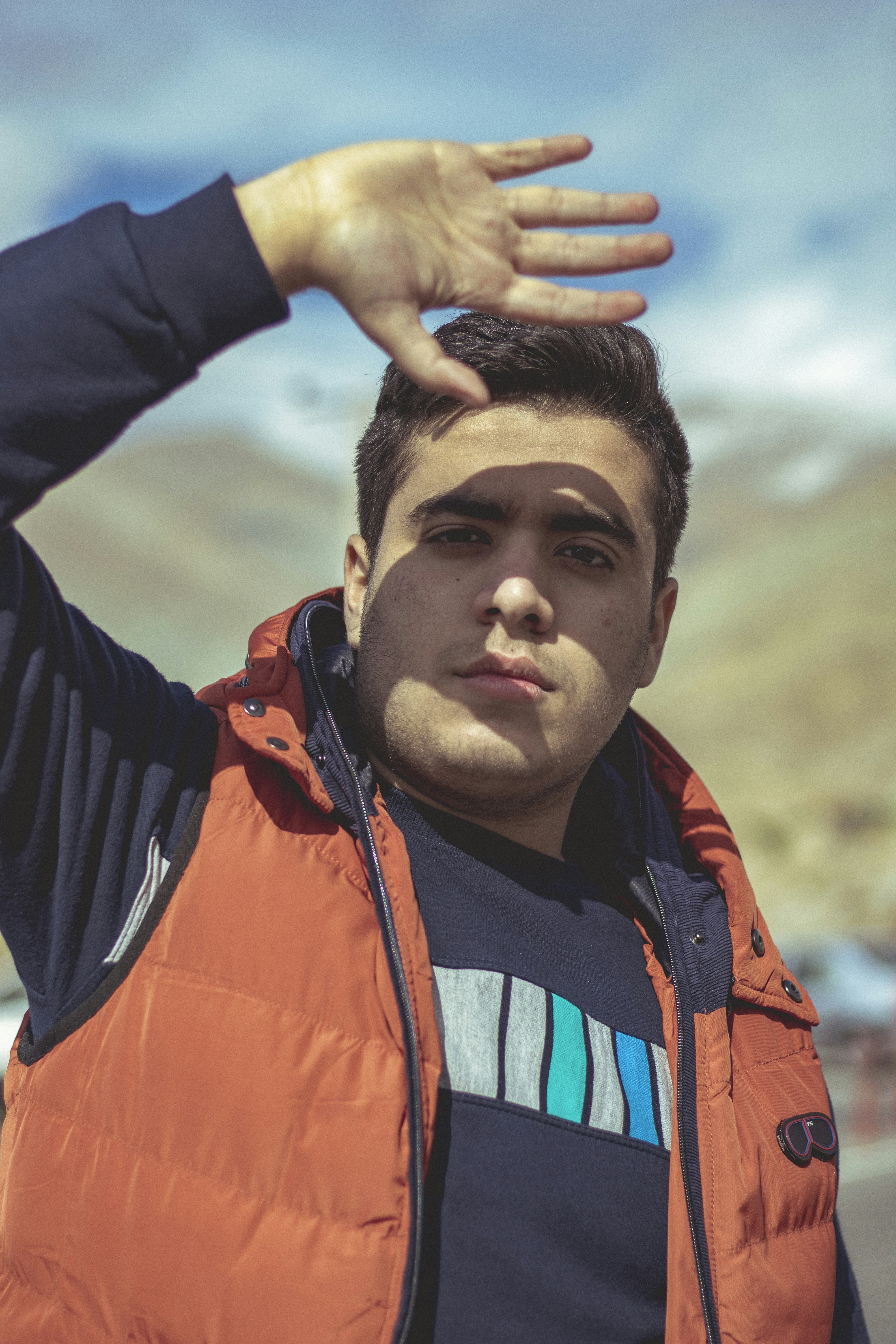 Young man shielding his eyes from the sun with a mountainous backdrop. His expression conveys a mix of contemplation and confidence.