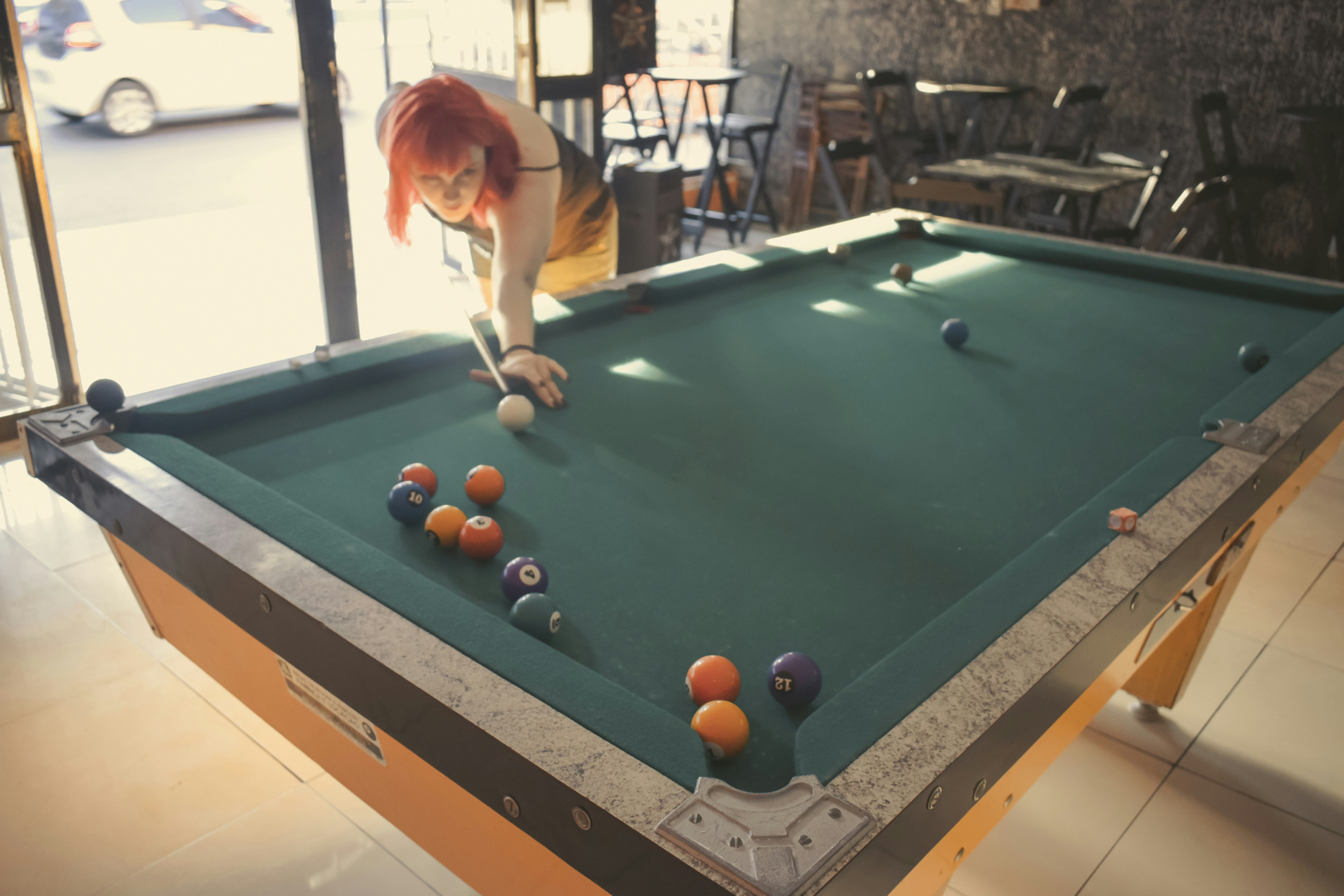 A player with vibrant red hair leans over a pool table, aiming for the cue ball amidst a colorful array of balls. The scene captures the intensity and strategy of billiards.