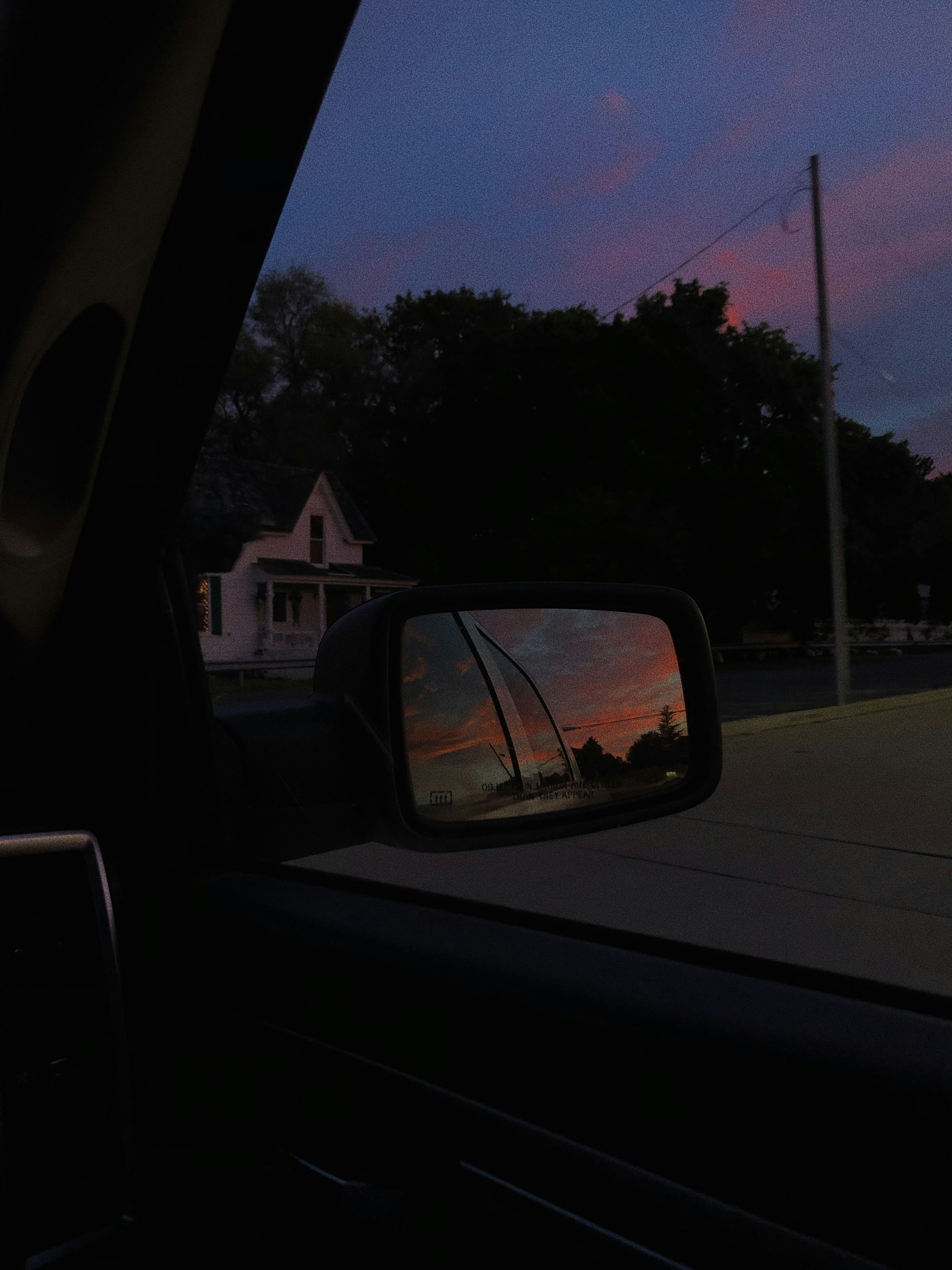 Side mirror reflecting a vibrant sunset with silhouettes of trees and a house in the background.