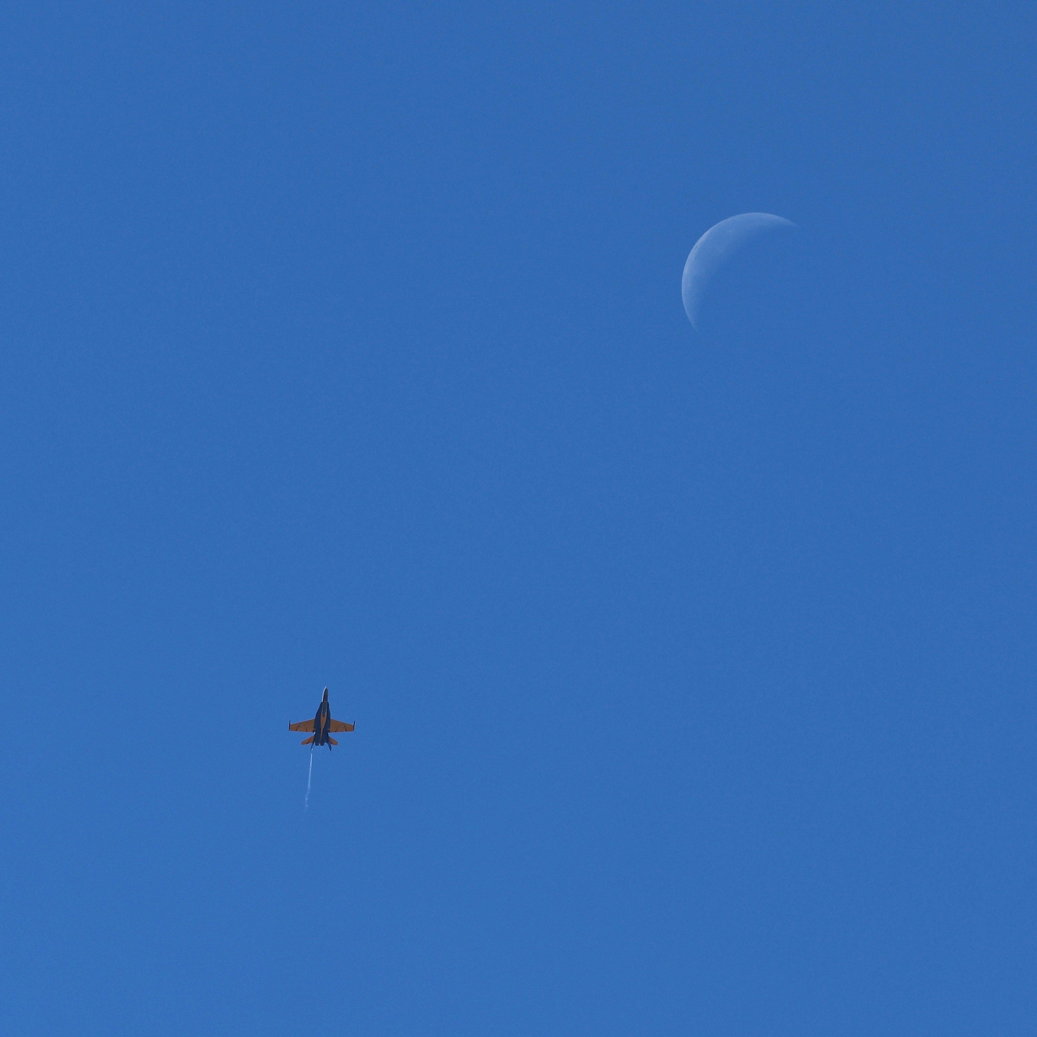 Jet against a clear blue dome under a pale crescent