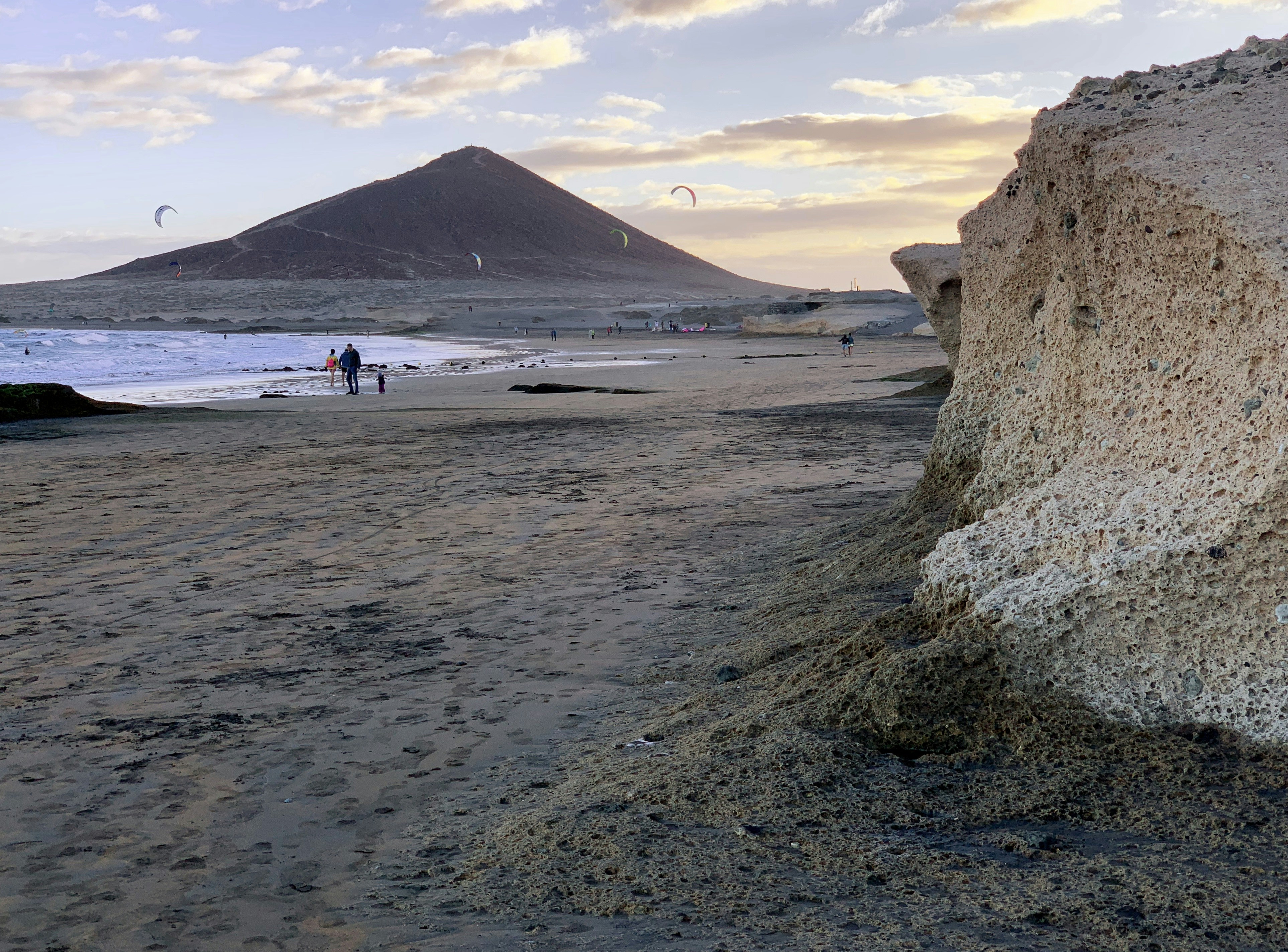 People walking on a sandy beach near a towering volcanic hill under a cloudy sky.
