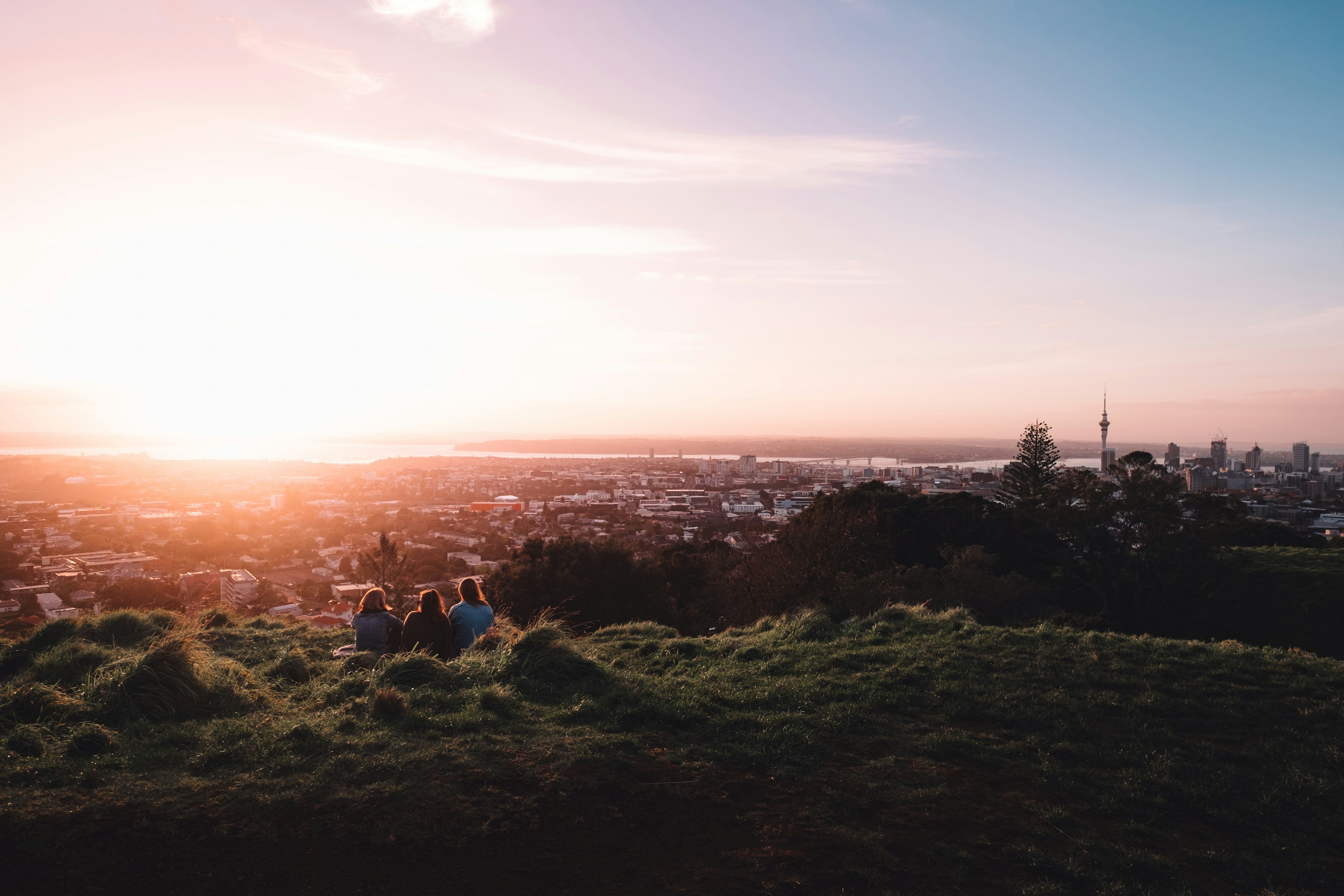 Three people sit on a grassy hill overlooking a city skyline under a vibrant sunset sky.
