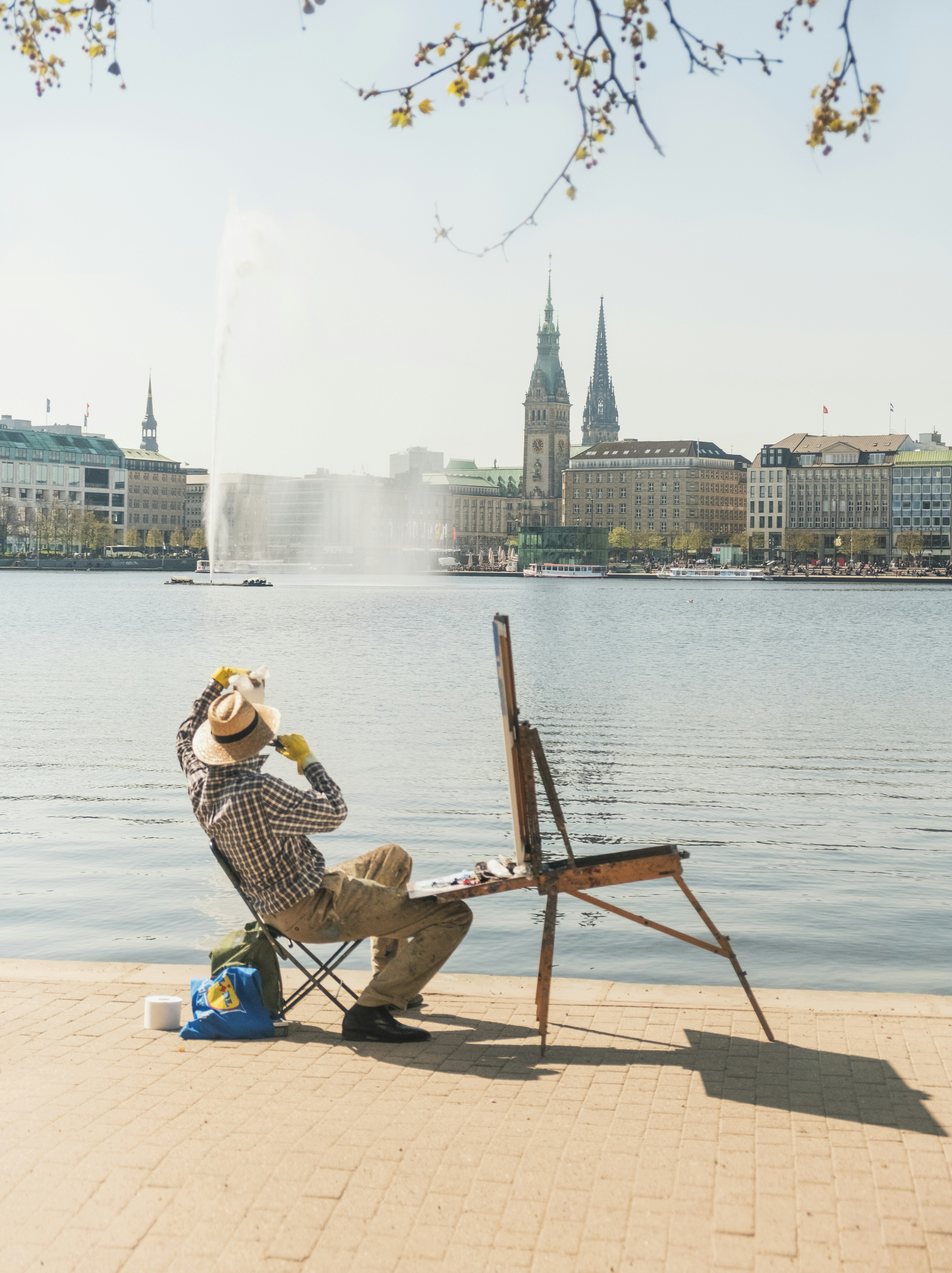 An artist captures the serene lakeside view, with historic buildings and a fountain in the background. The scene conveys a peaceful moment of creativity.